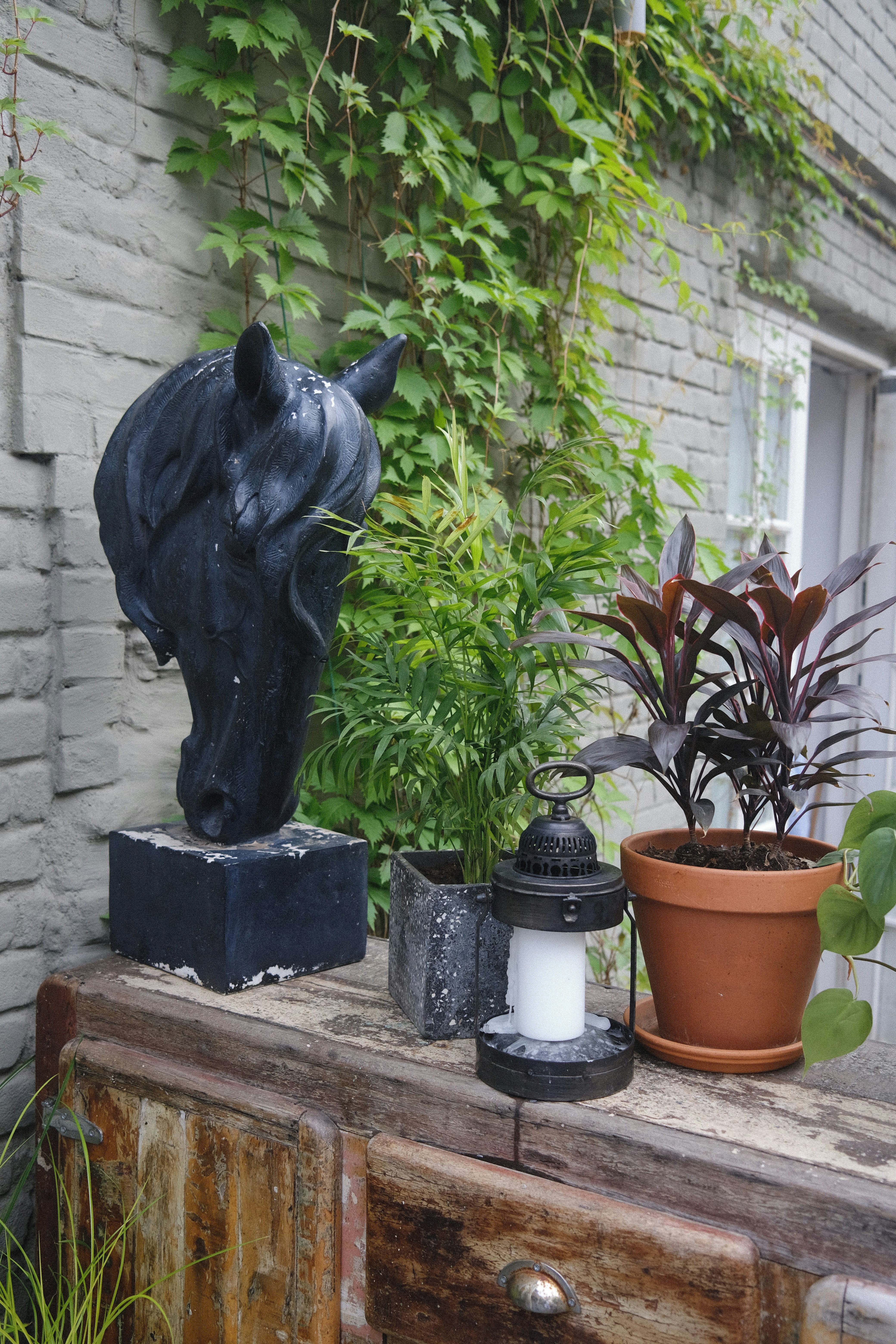 Dark horsehead sculpture guards a sunlit patio garden with lantern and potted greenery
