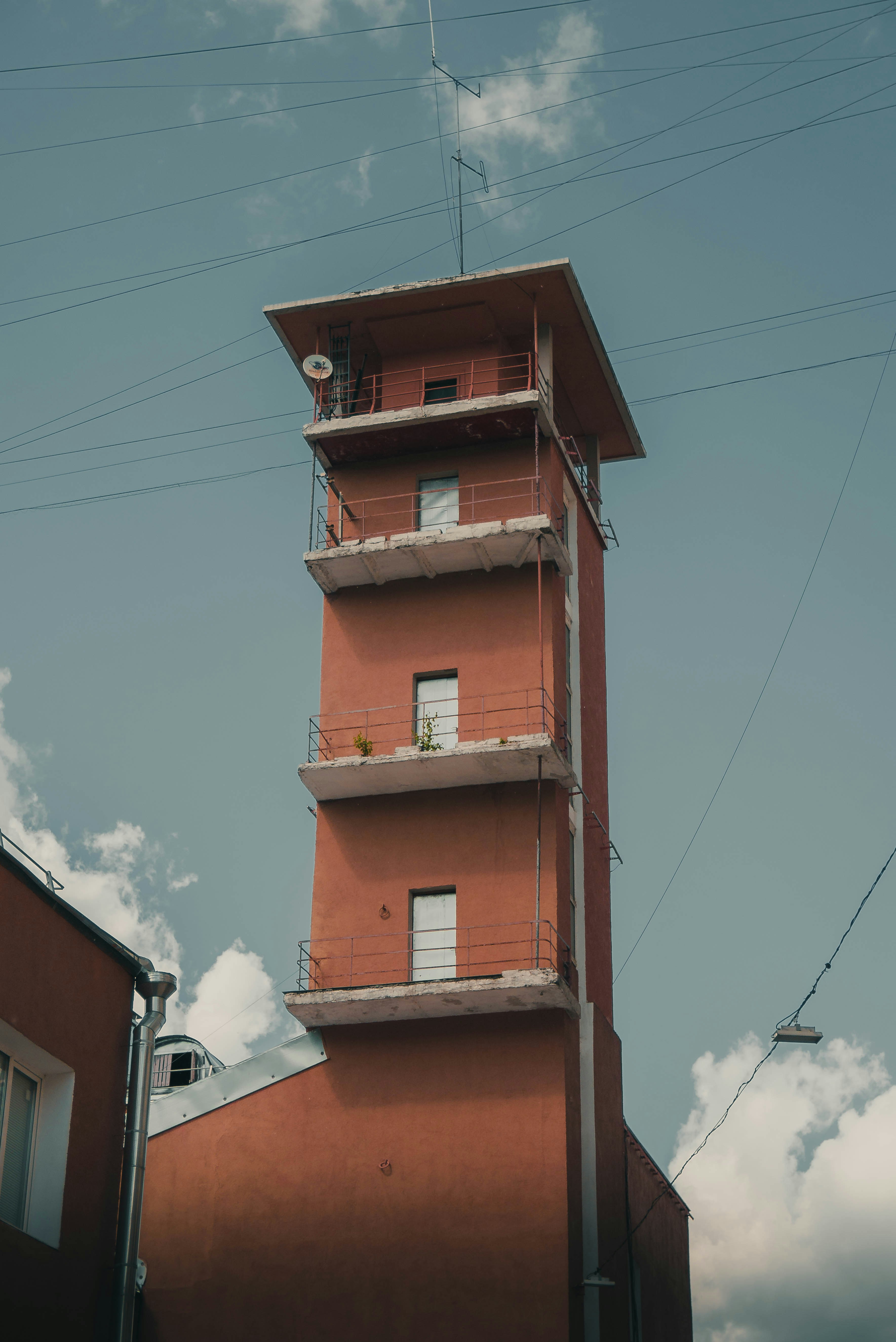 A tall, orange tower with balconies and windows, set against a backdrop of a blue sky with scattered clouds. The structure features antennas and power lines.