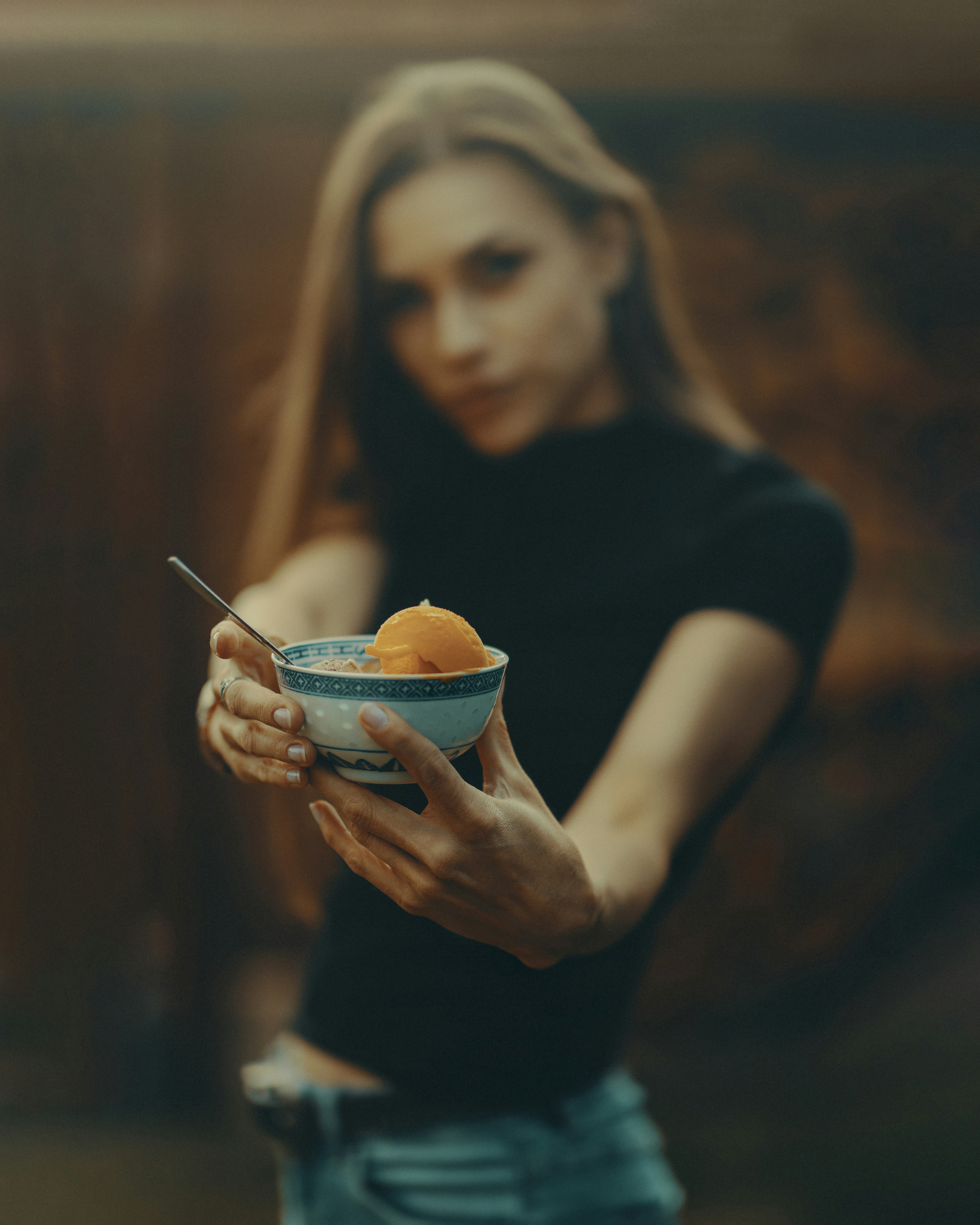 A young woman holds a bowl of bright orange sorbet with a spoon, showcasing a moment of indulgence. The background is softly blurred, emphasizing the vibrant dessert.