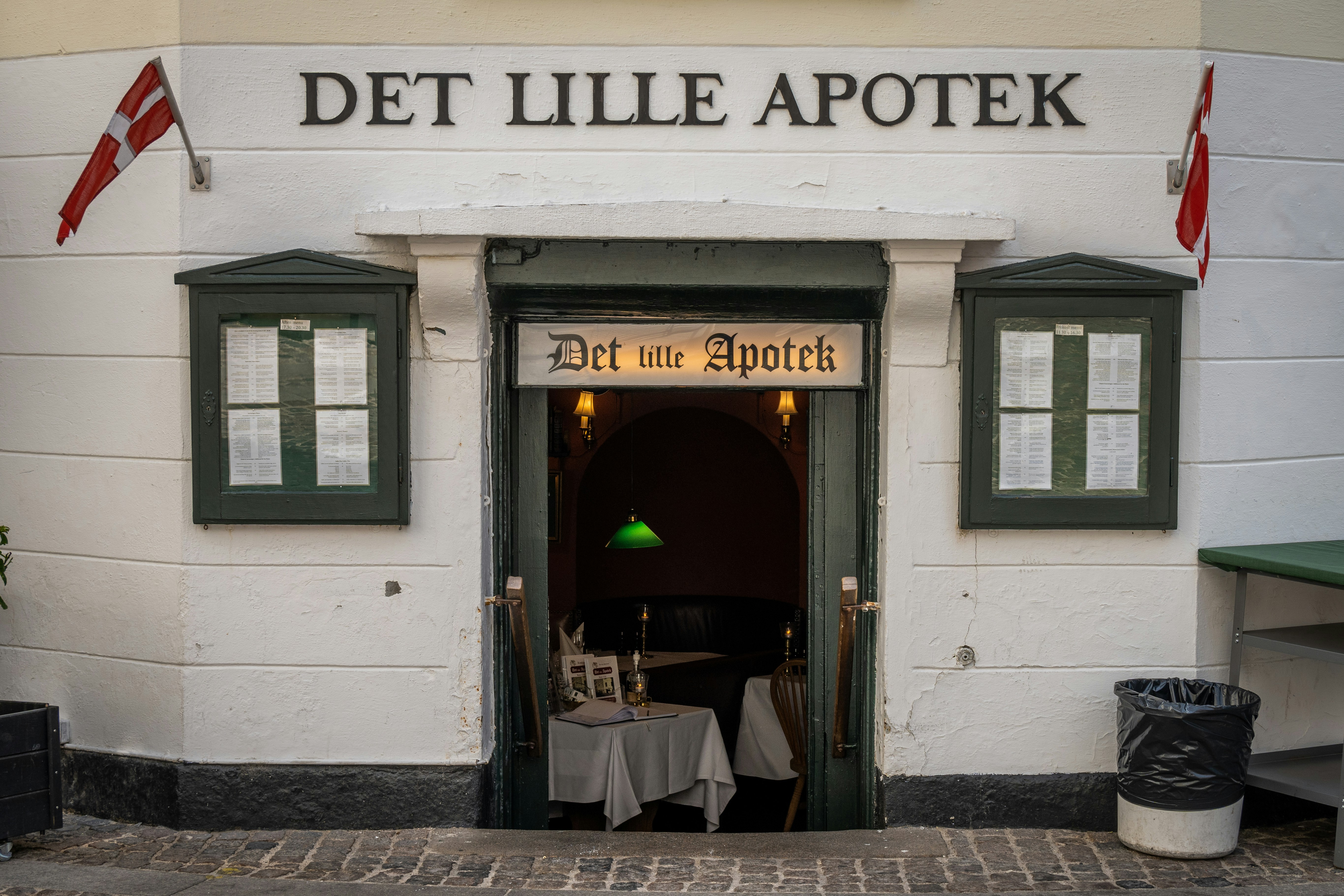 Charming entrance of Det Lille Apotek, featuring green shutters and a welcoming glow from the interior. A glimpse into Danish dining culture.
