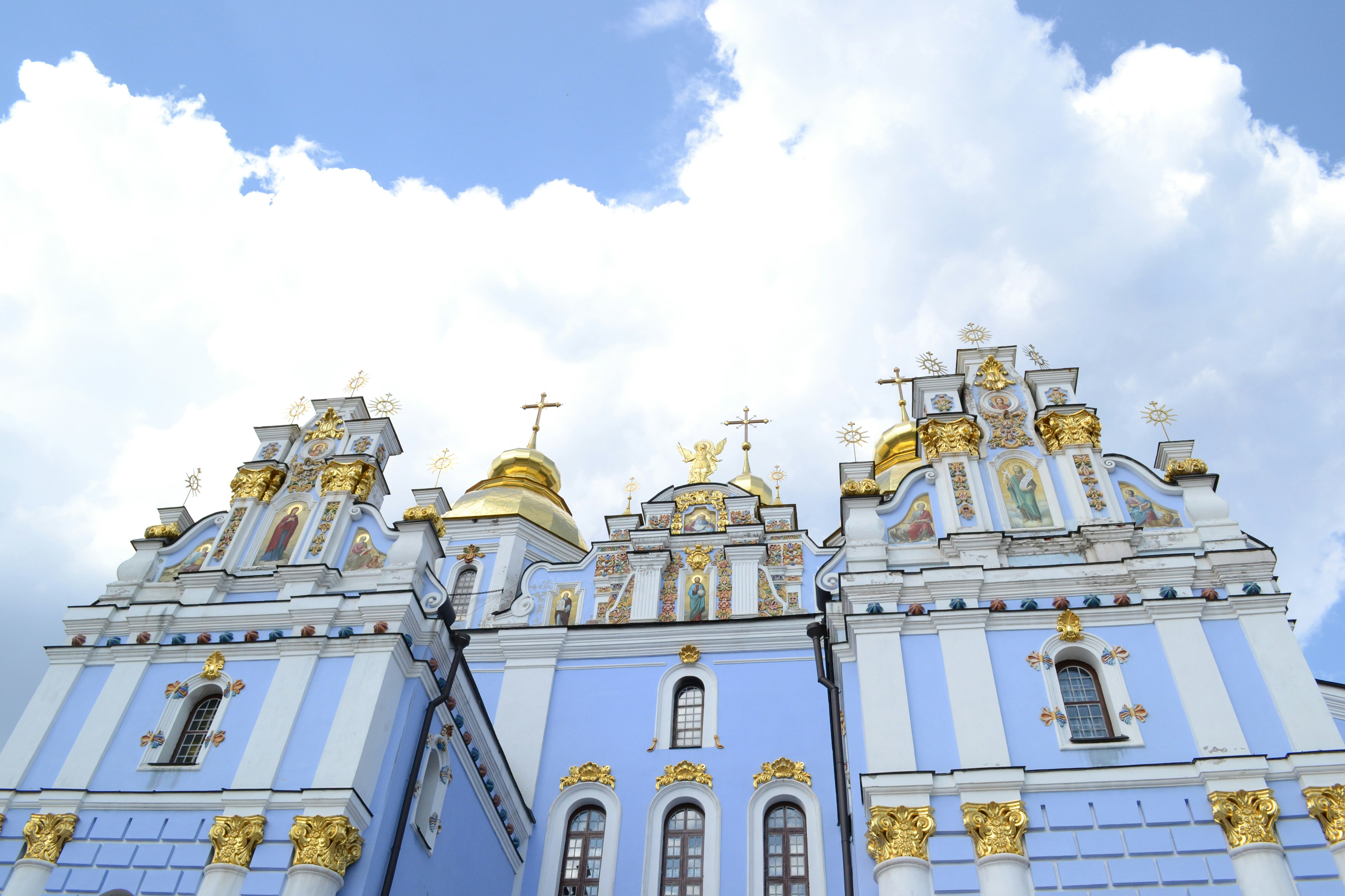 Ornate church facade with blue and white detailing, crowned by golden domes beneath a bright sky.
