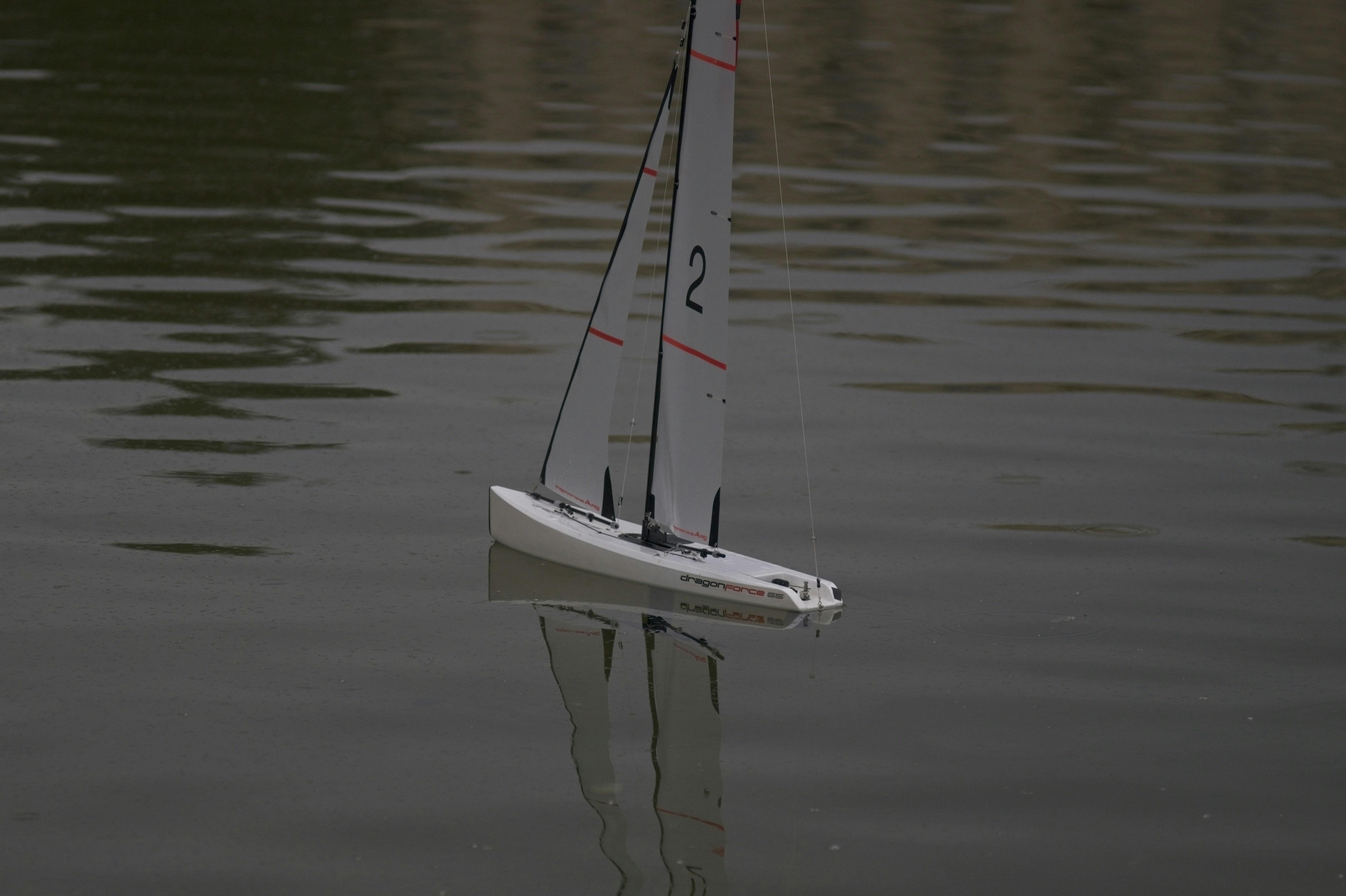 A model sailboat glides smoothly across a calm body of water, its reflection mirrored perfectly on the surface. The number '2' is visible on the sail.