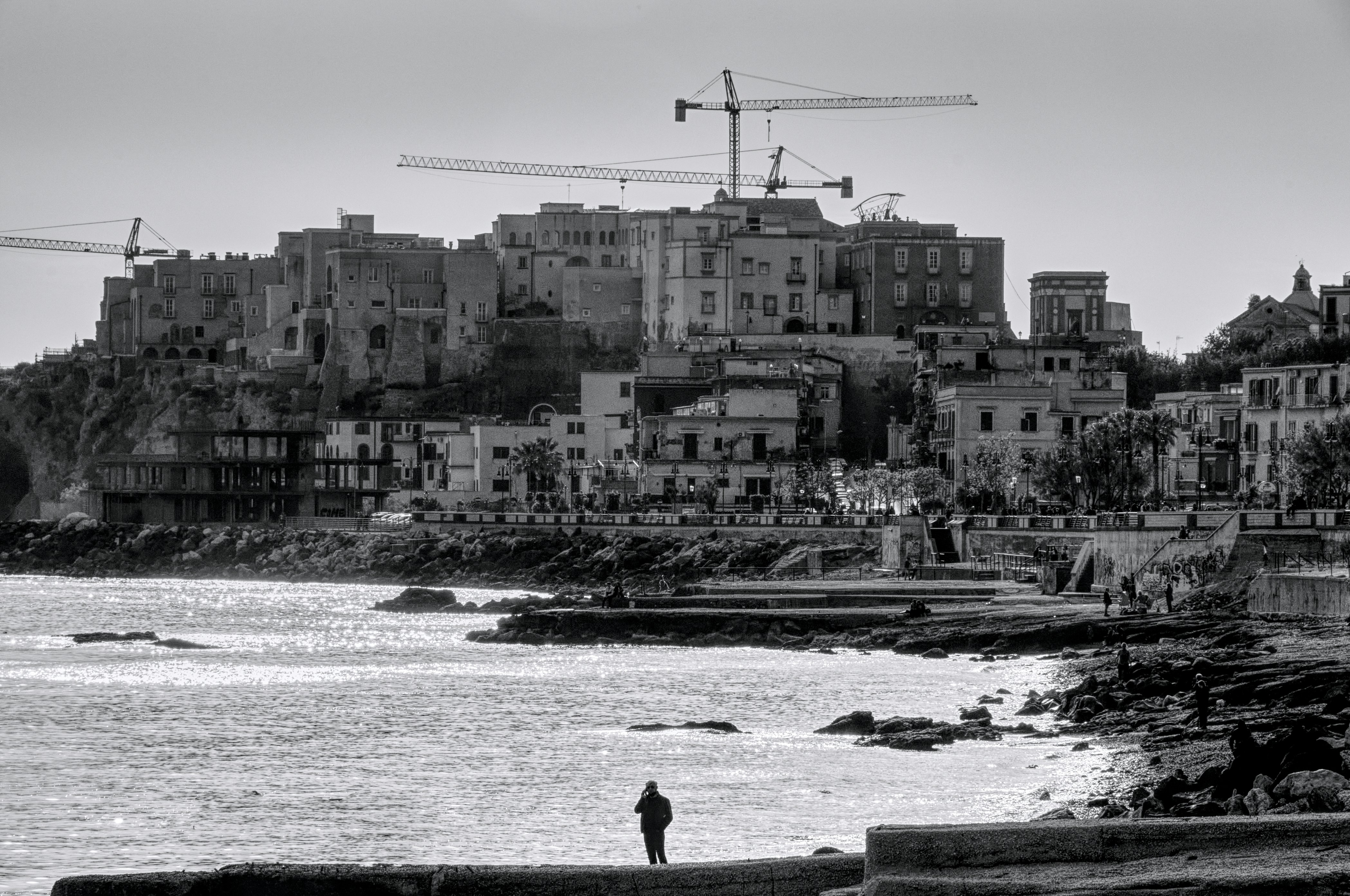 Monochrome scene of a lone figure walking by the seaside with urban buildings and construction cranes in the background.