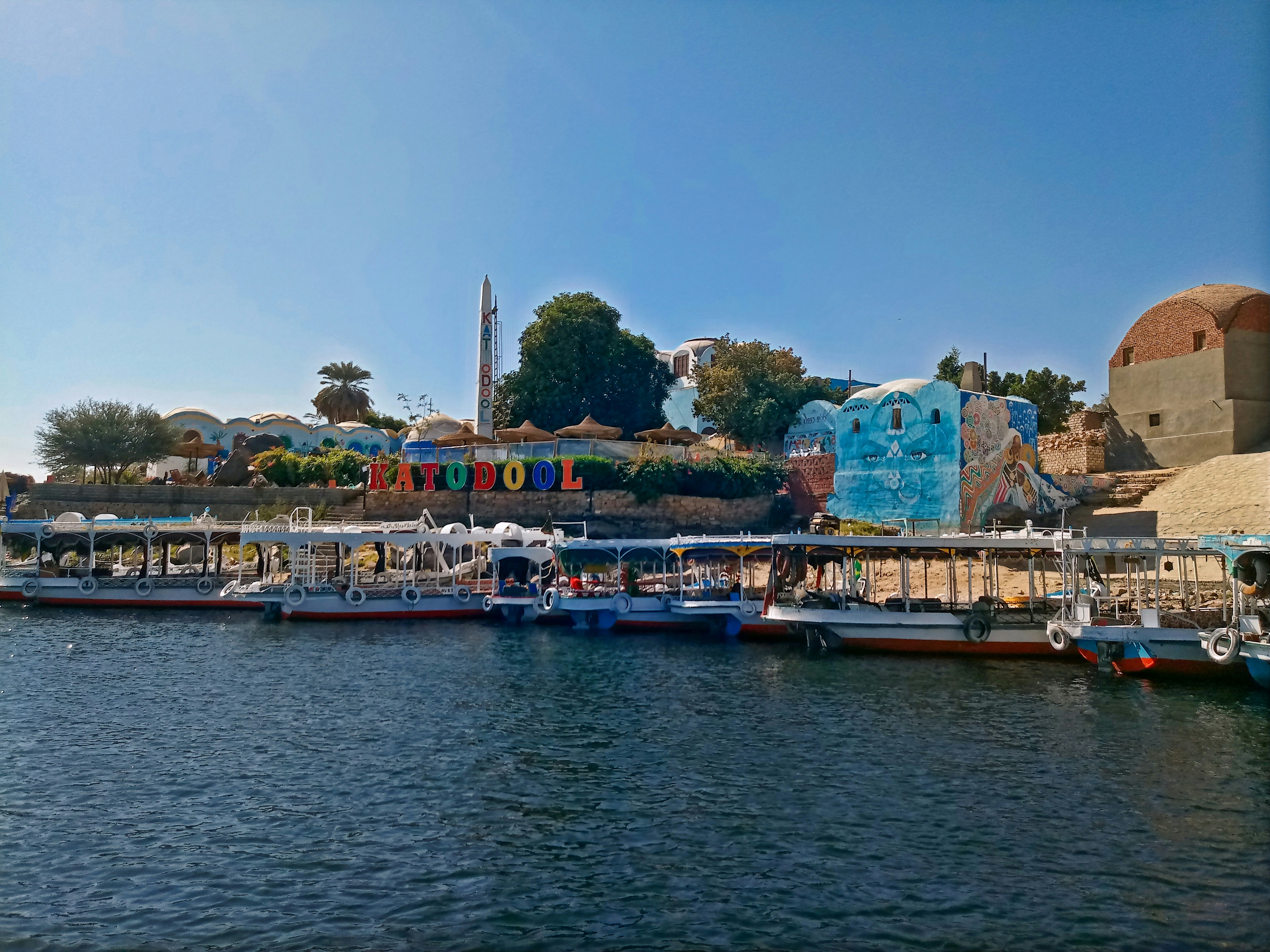 Colorful buildings line the waterfront of a Nubian village along the Nile River under a clear blue sky.
