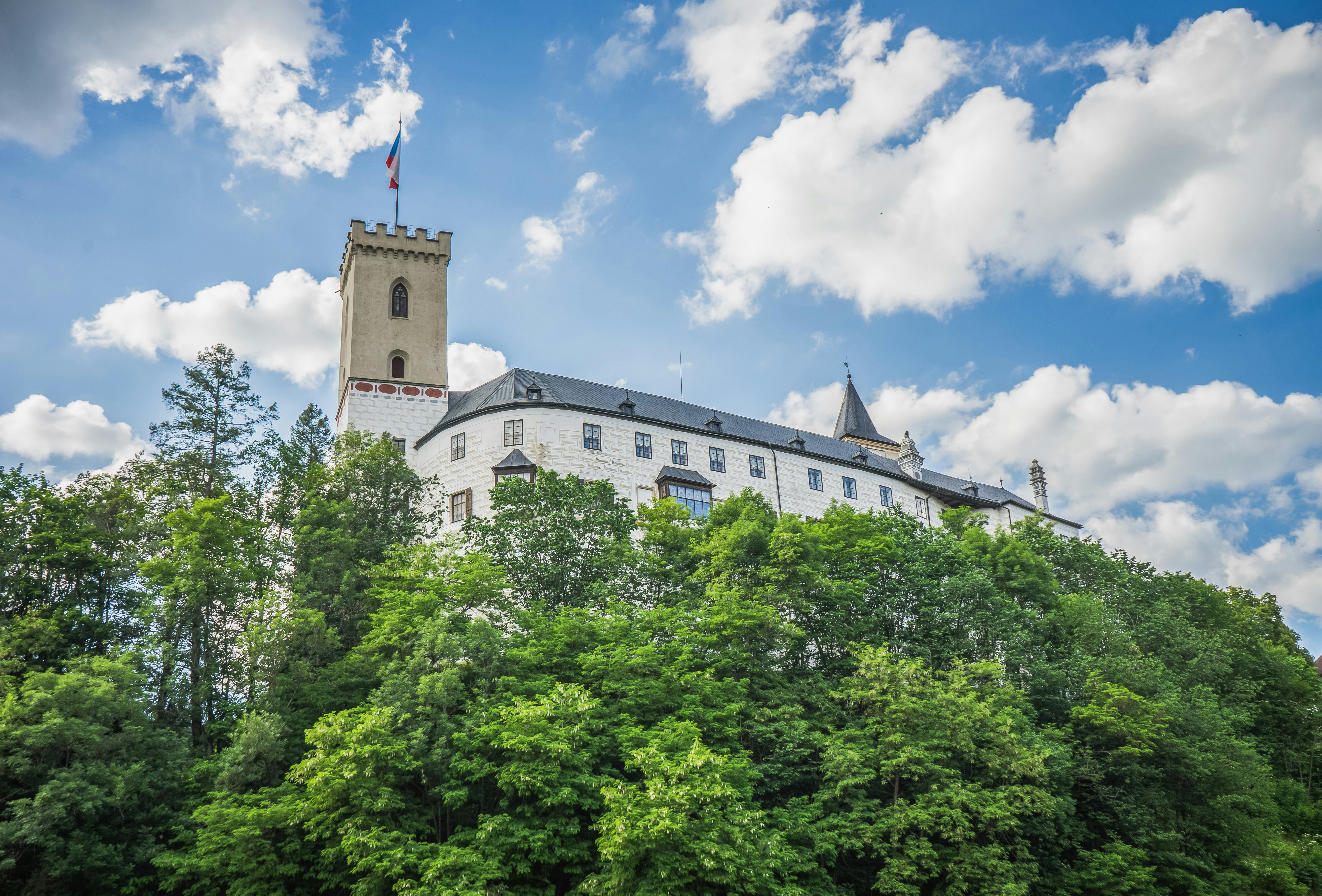 Historic castle rising above lush green trees against a backdrop of a cloudy blue sky.