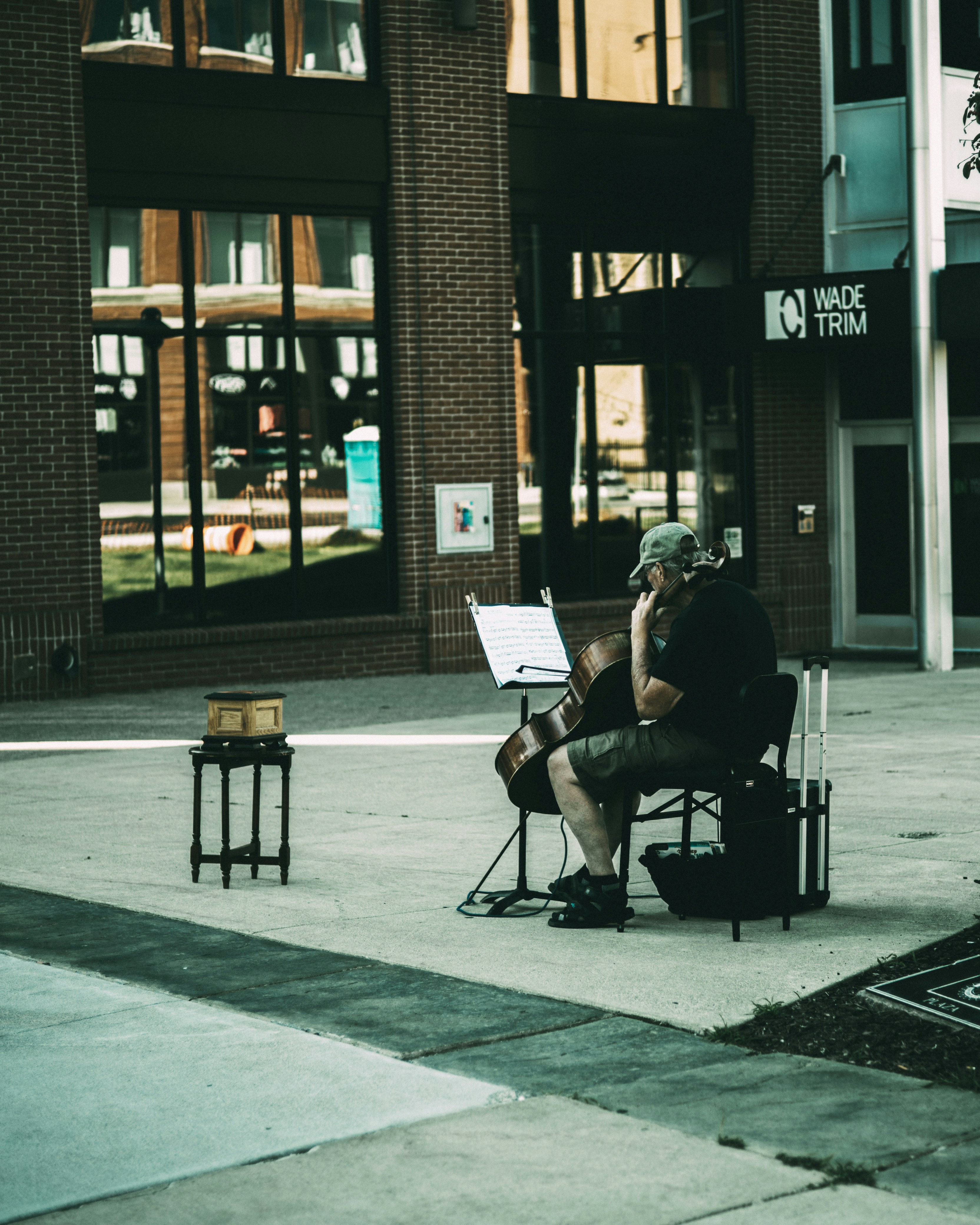 Musician seated on the pavement, immersed in practice with a cello, accompanied by a small stand and a wooden box. Reflections of buildings create a dynamic urban backdrop.
