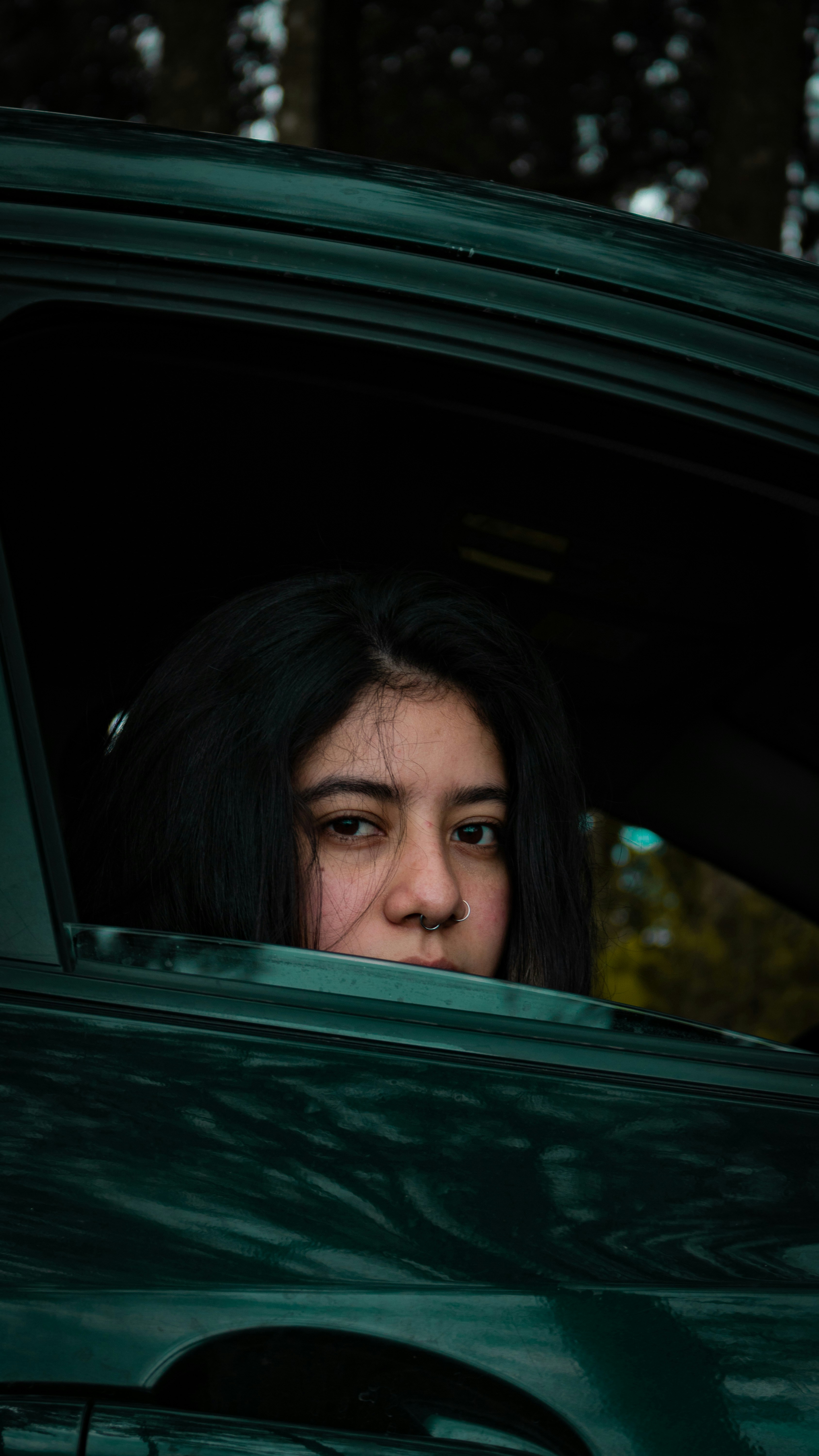 A young woman gazes thoughtfully from a car window, framed by dark green hues. Her expression invites curiosity and reflection.