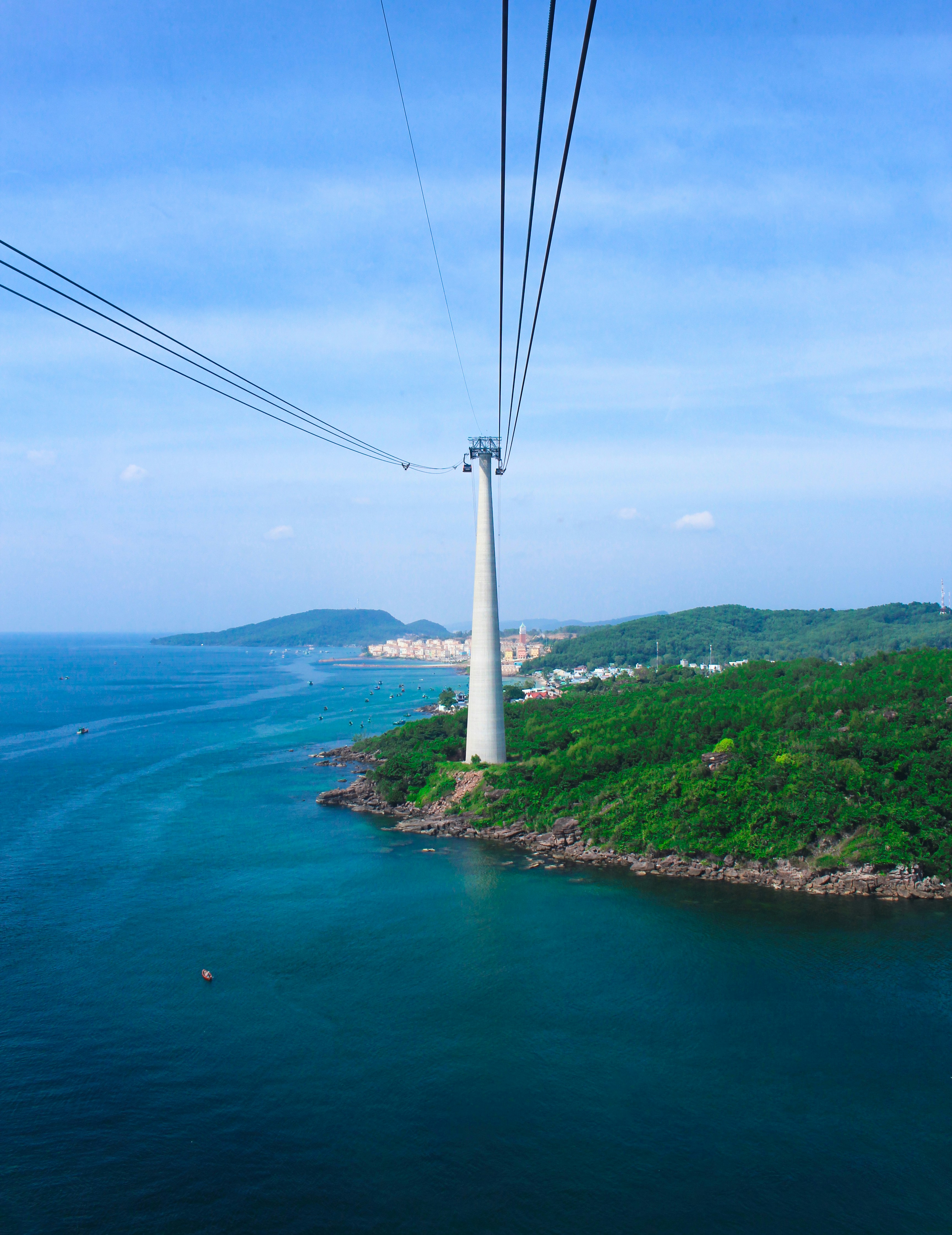 A cableway tower stands tall above the turquoise waters, connecting lush green hills with the distant coastline. The scene captures the harmony of nature and human engineering.
