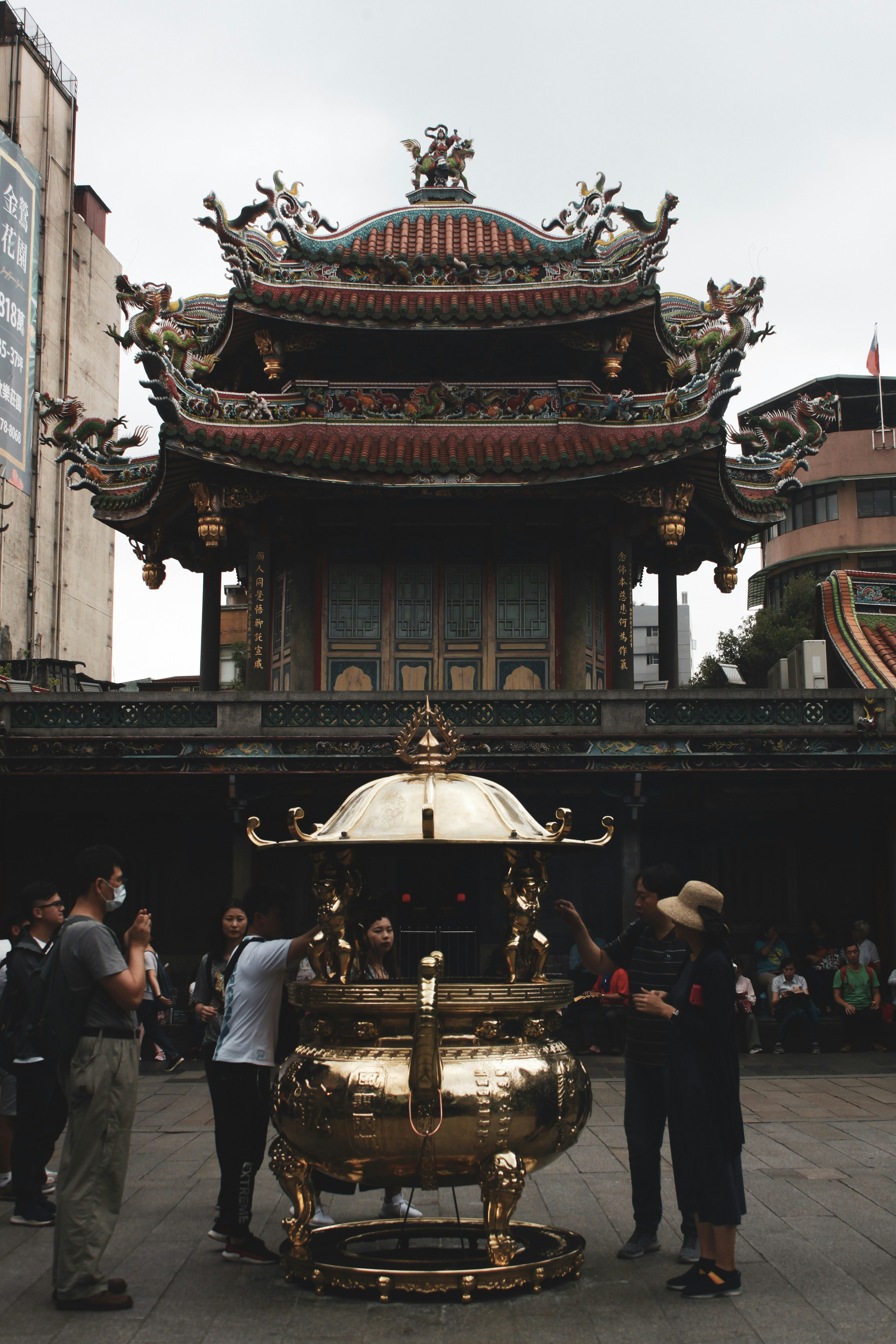 Visitors engage in a cultural ritual around a large, ornate golden incense burner in front of a traditional temple.