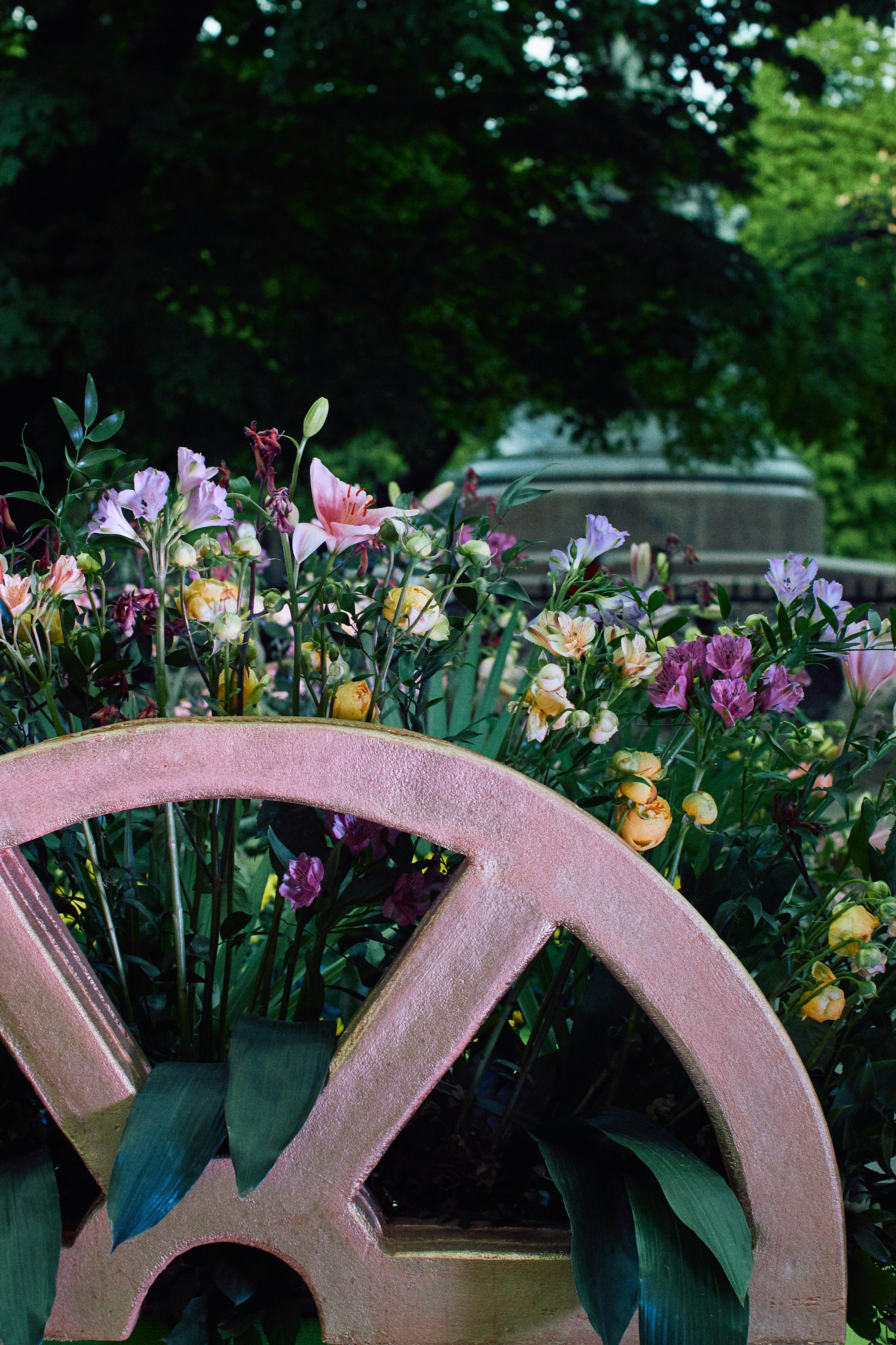 A delicate arrangement of colorful flowers blooms around a rustic stone wheel, creating a serene garden scene. The lush greenery in the background enhances the vibrant colors of the flowers.