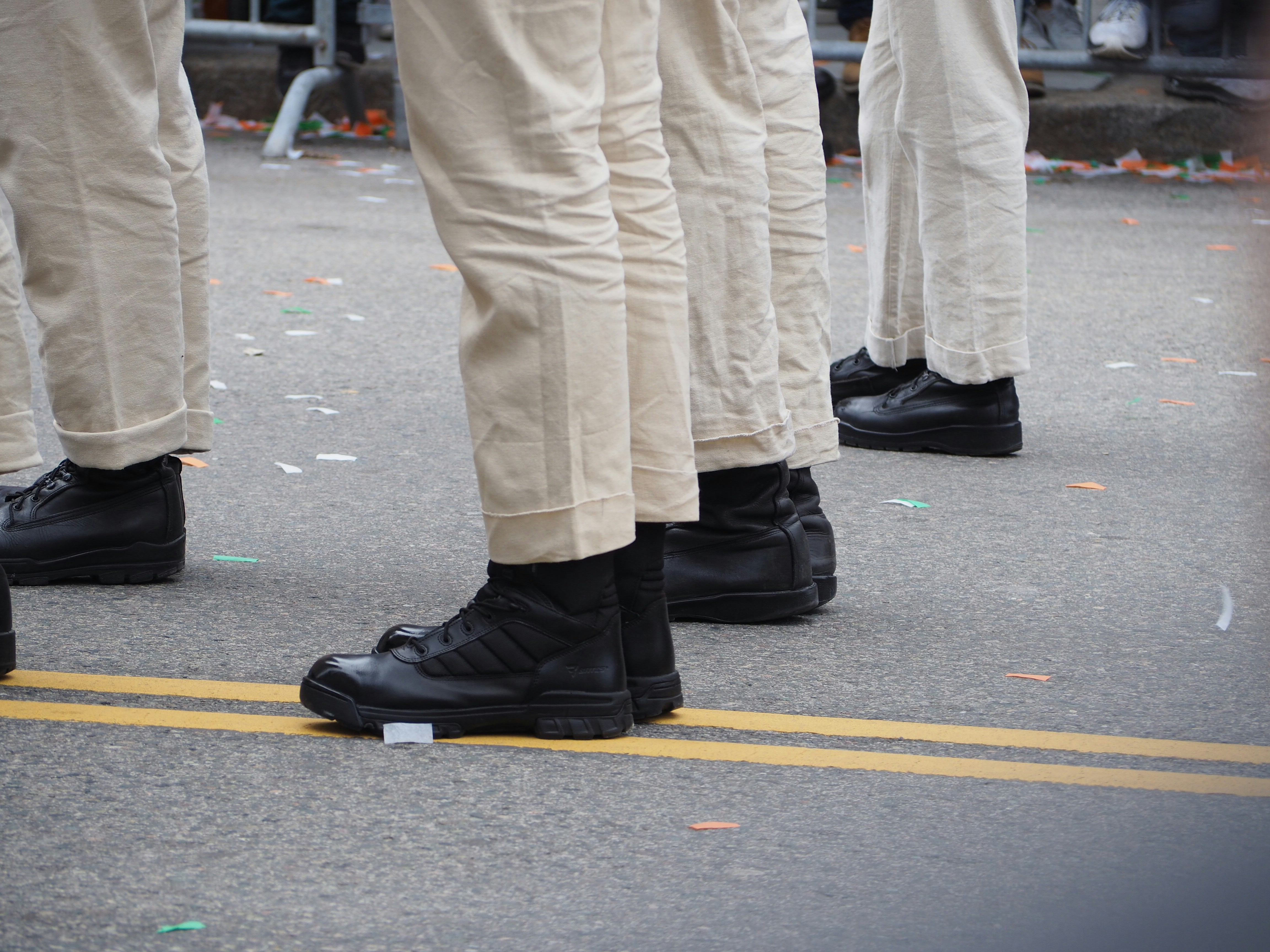 Close-up of uniformed feet in black boots standing in formation on a parade route, highlighting the discipline and precision of the participants.