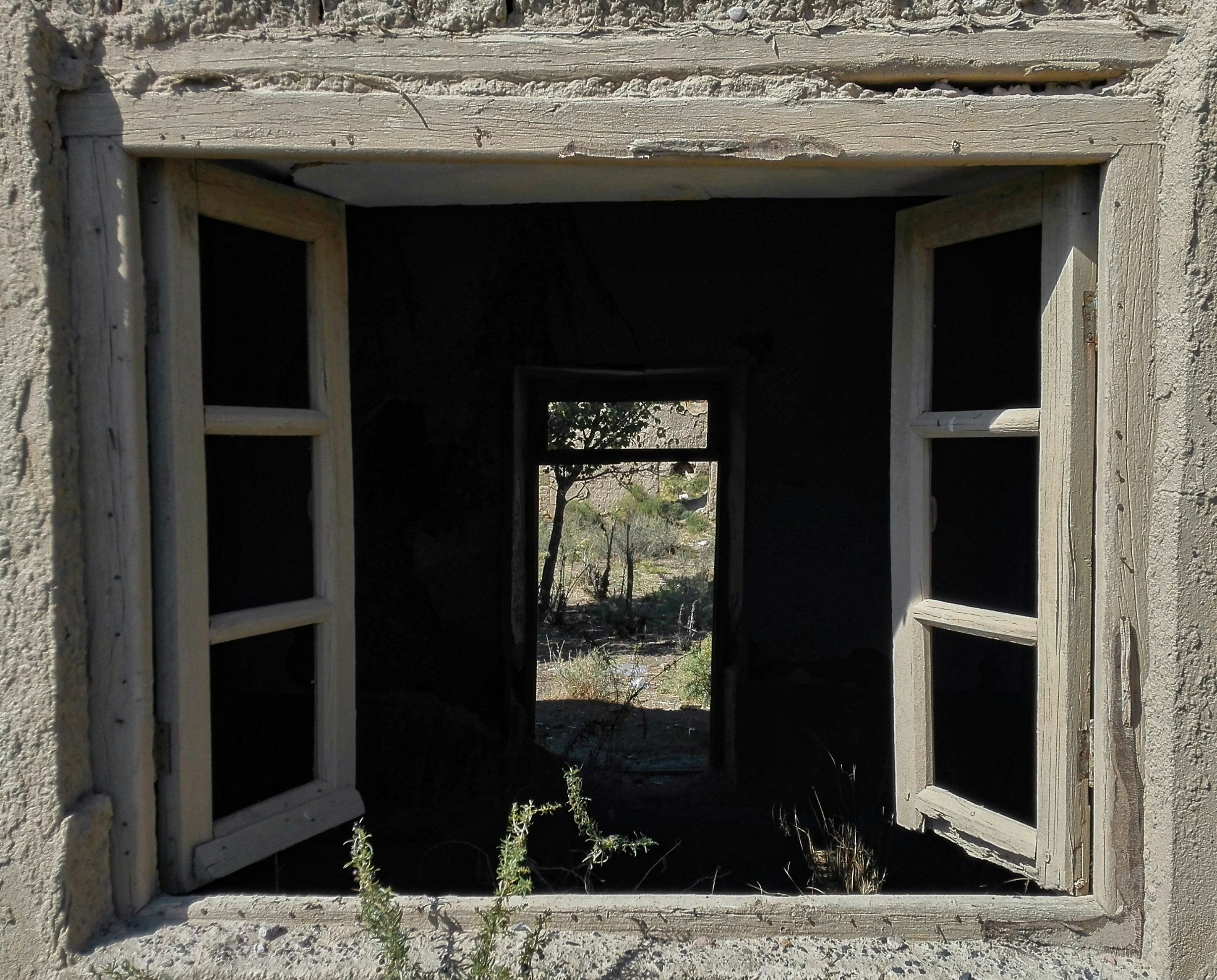 View through an open window into a dark room, revealing another window and overgrown greenery beyond. The scene evokes a sense of abandonment and nostalgia.