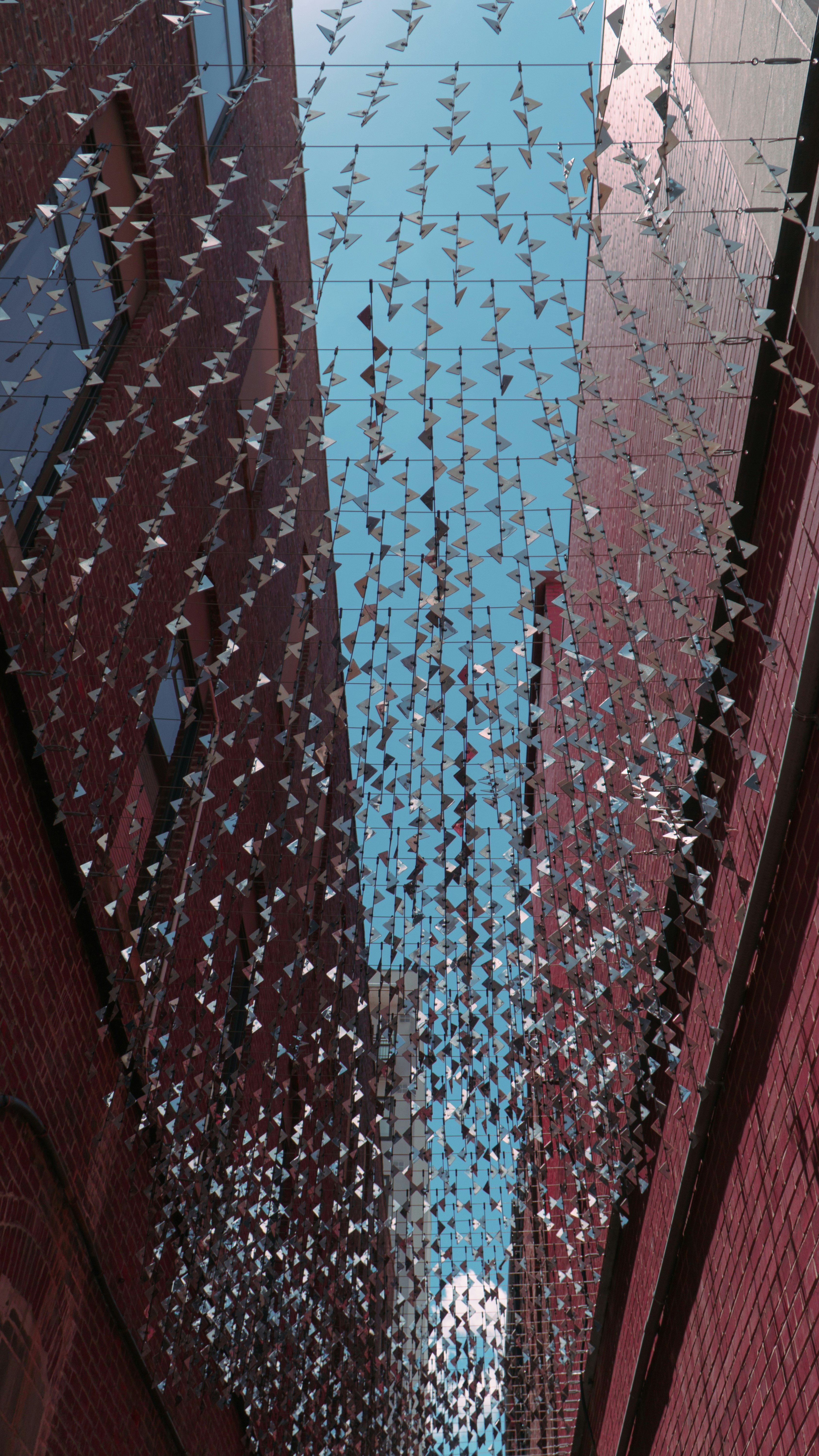 Mirrored paper cranes suspended in an urban alley, creating a whimsical canopy against a bright blue sky.