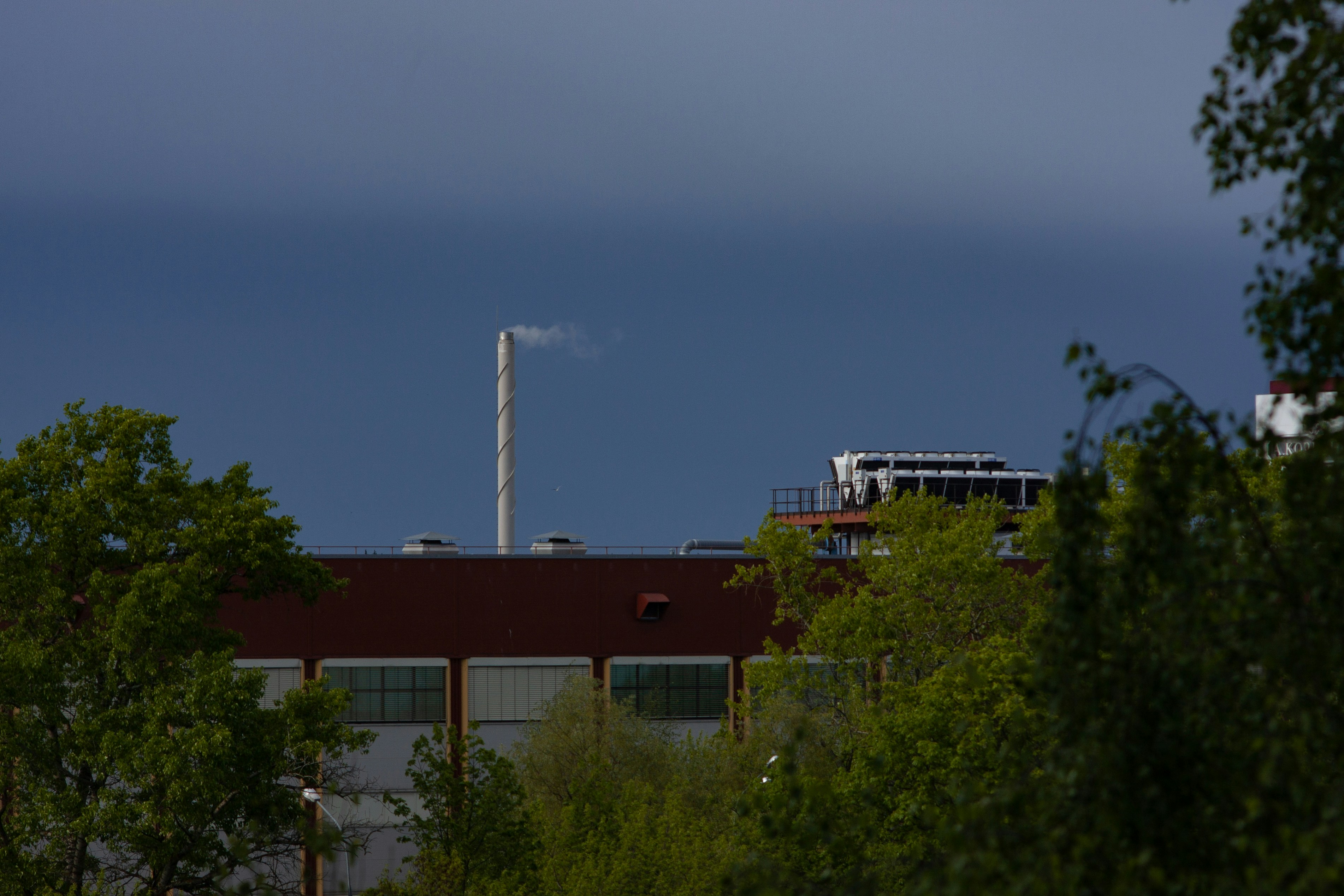 A factory's smokestack rises against a backdrop of lush greenery and a moody sky, illustrating the juxtaposition of industrial and natural elements.