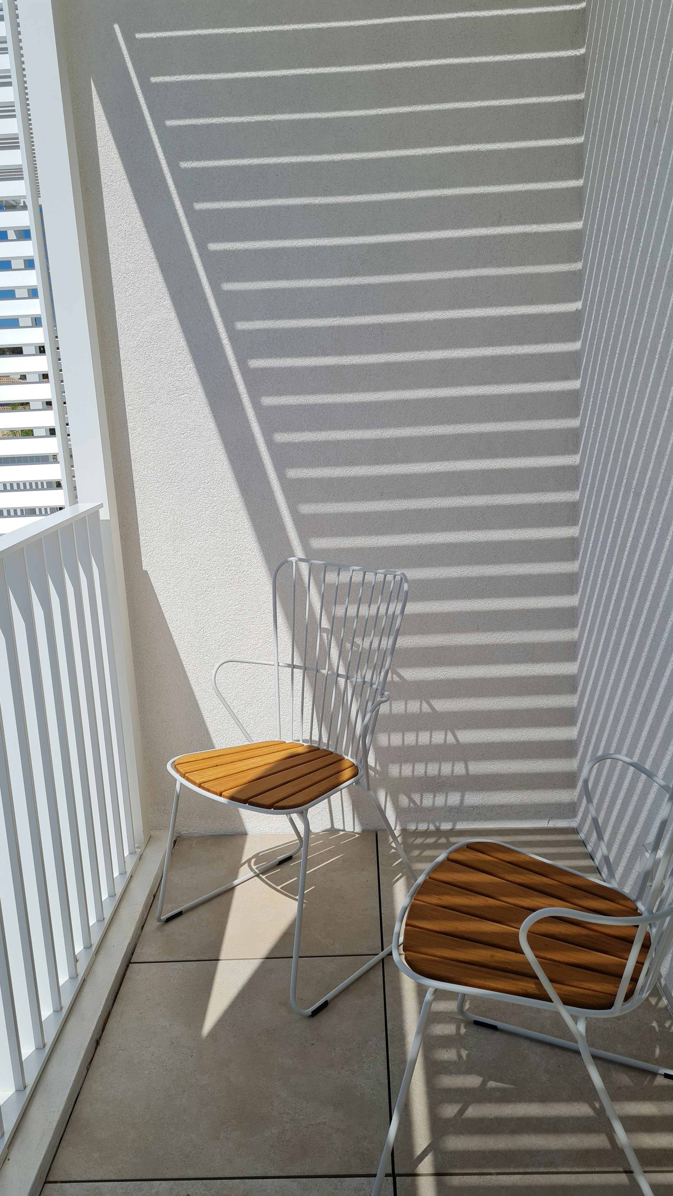 Two modern chairs with wooden seats cast intricate shadows against a bright wall, creating a play of light and geometry.