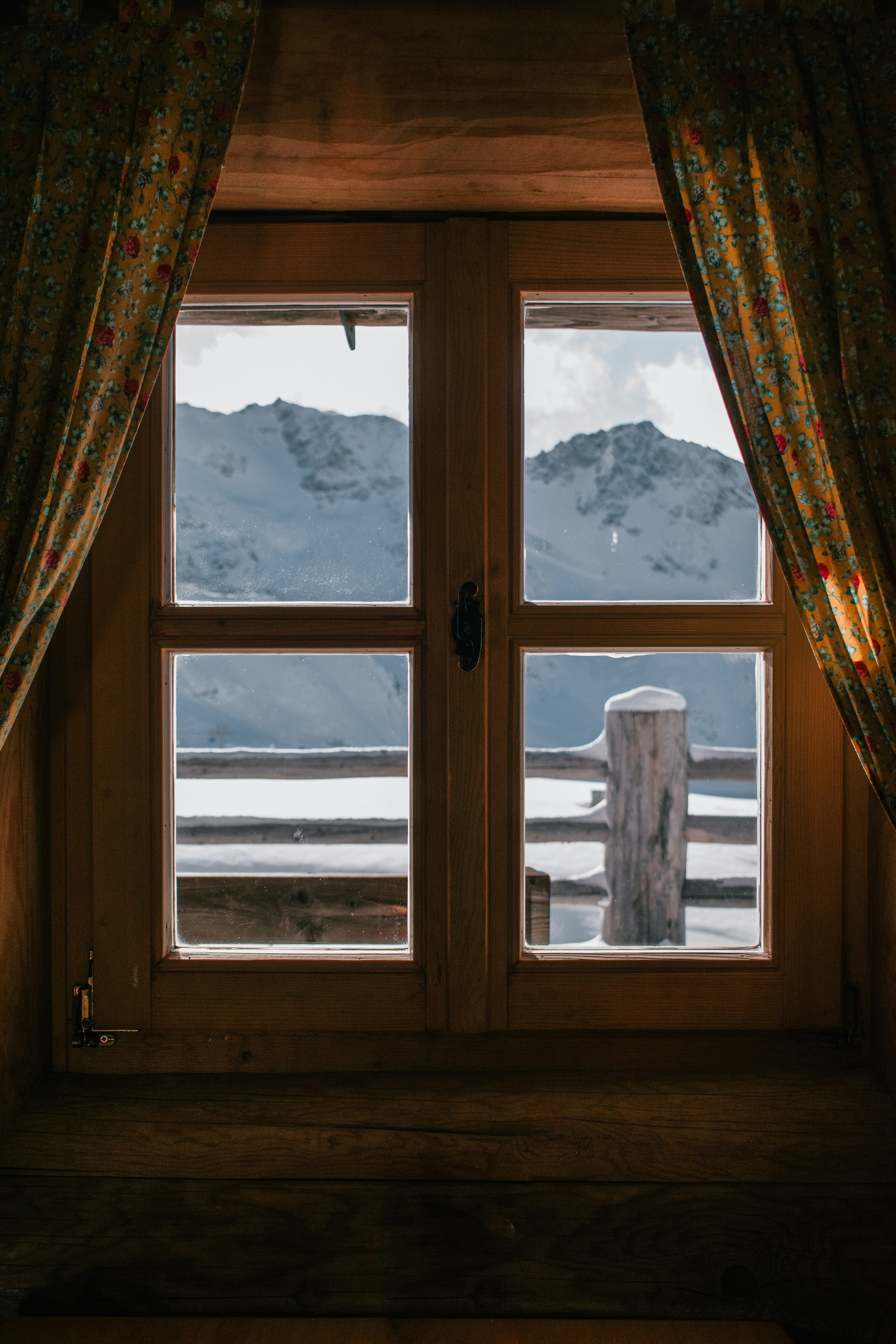Wooden window adorned with floral curtains, framing a snowy mountain landscape beyond. The scene evokes a sense of warmth and tranquility.