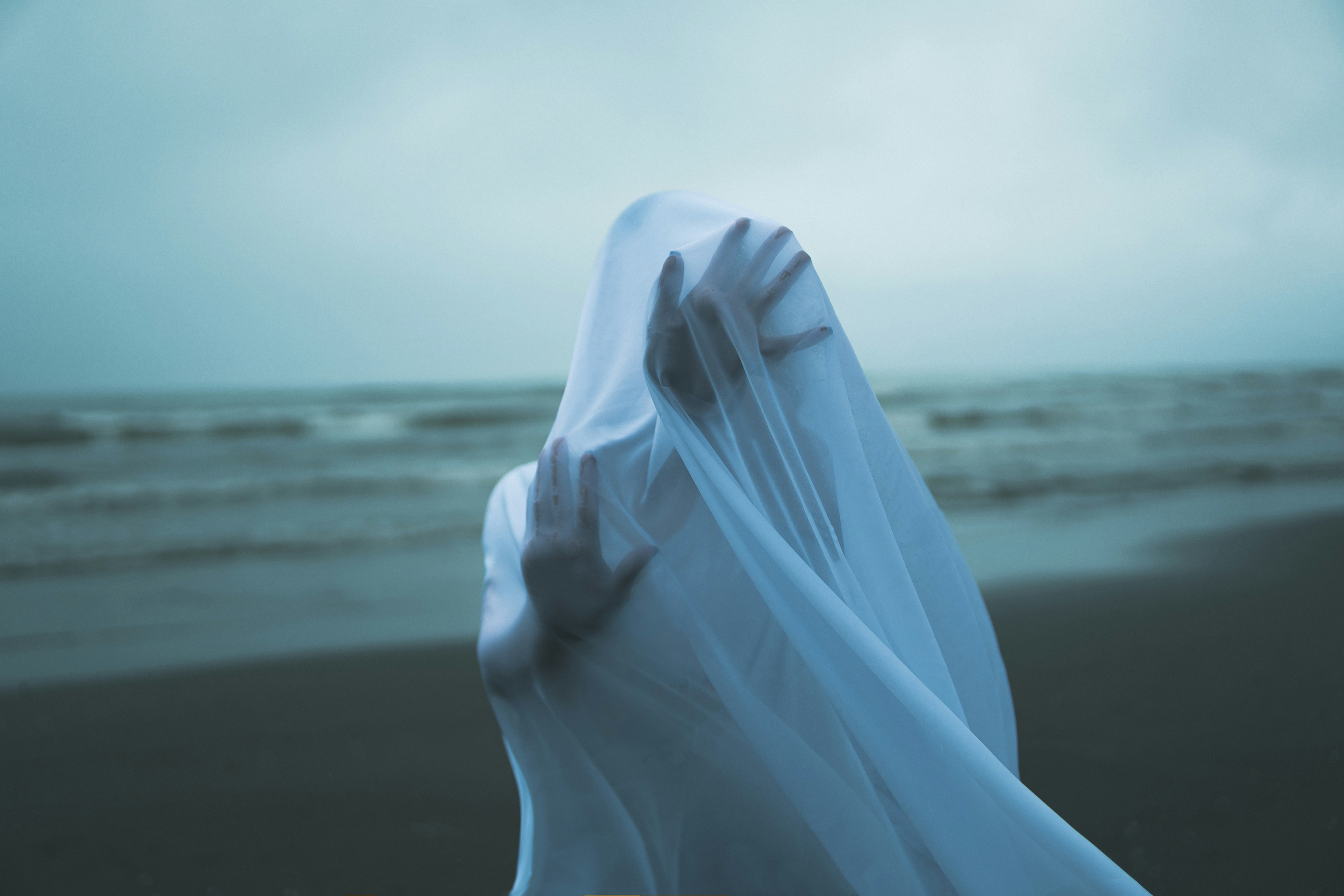 This mesmerizing image captures a figure shrouded in a translucent blue veil against the backdrop of a misty beach. The ethereal blue tones and soft lighting create a dreamlike atmosphere, with the fabric's delicate texture and the ocean's gentle waves enhancing the composition's mysterious allure. The interplay of light and shadow on the veil and the serene coastal setting make this image visually striking.