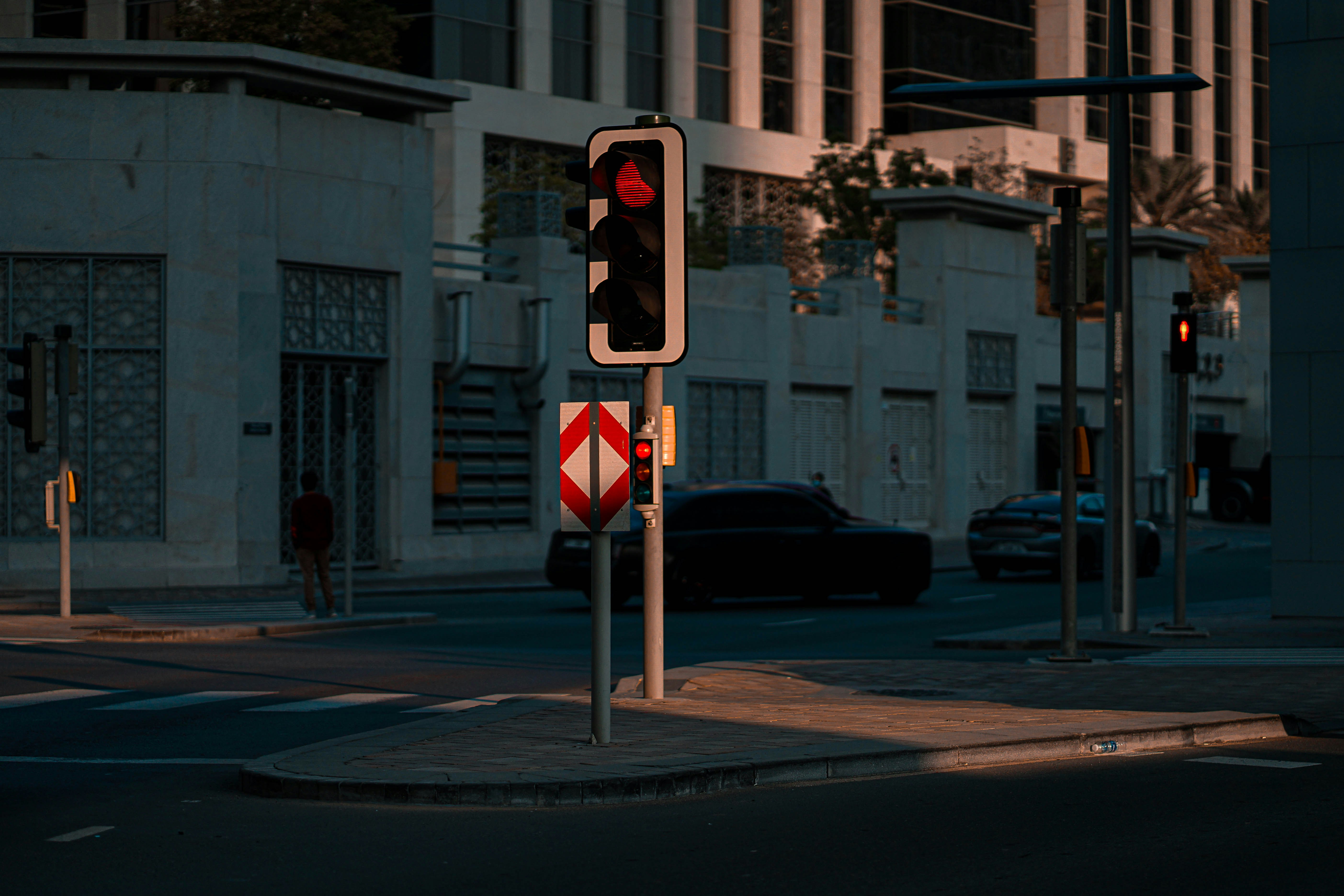Traffic light signaling red at a city intersection, with shadows cast by surrounding buildings during twilight.