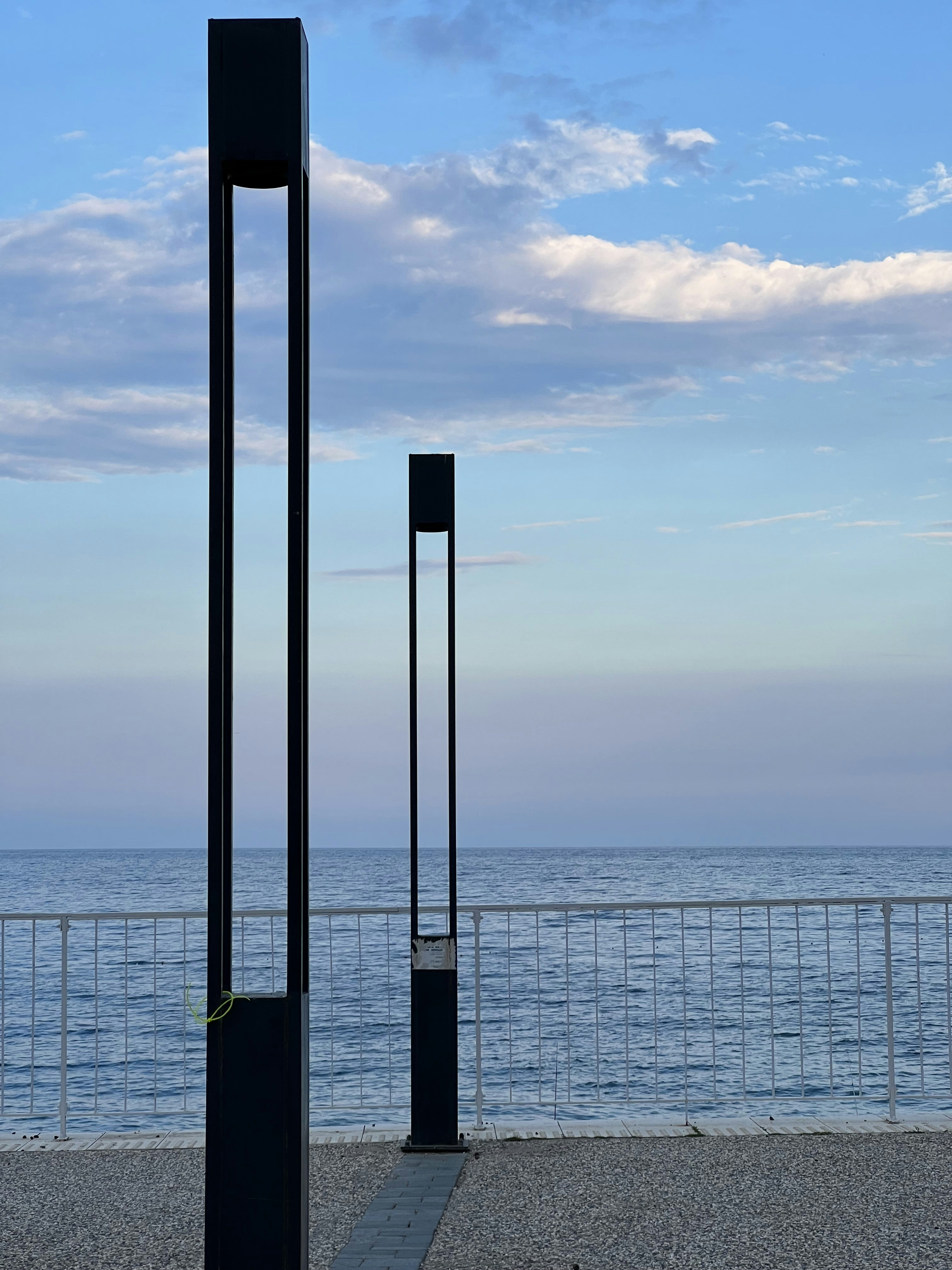 Two tall, slender sculptures stand against a serene ocean backdrop, framed by a soft blue sky. The scene captures the interplay between art and nature.