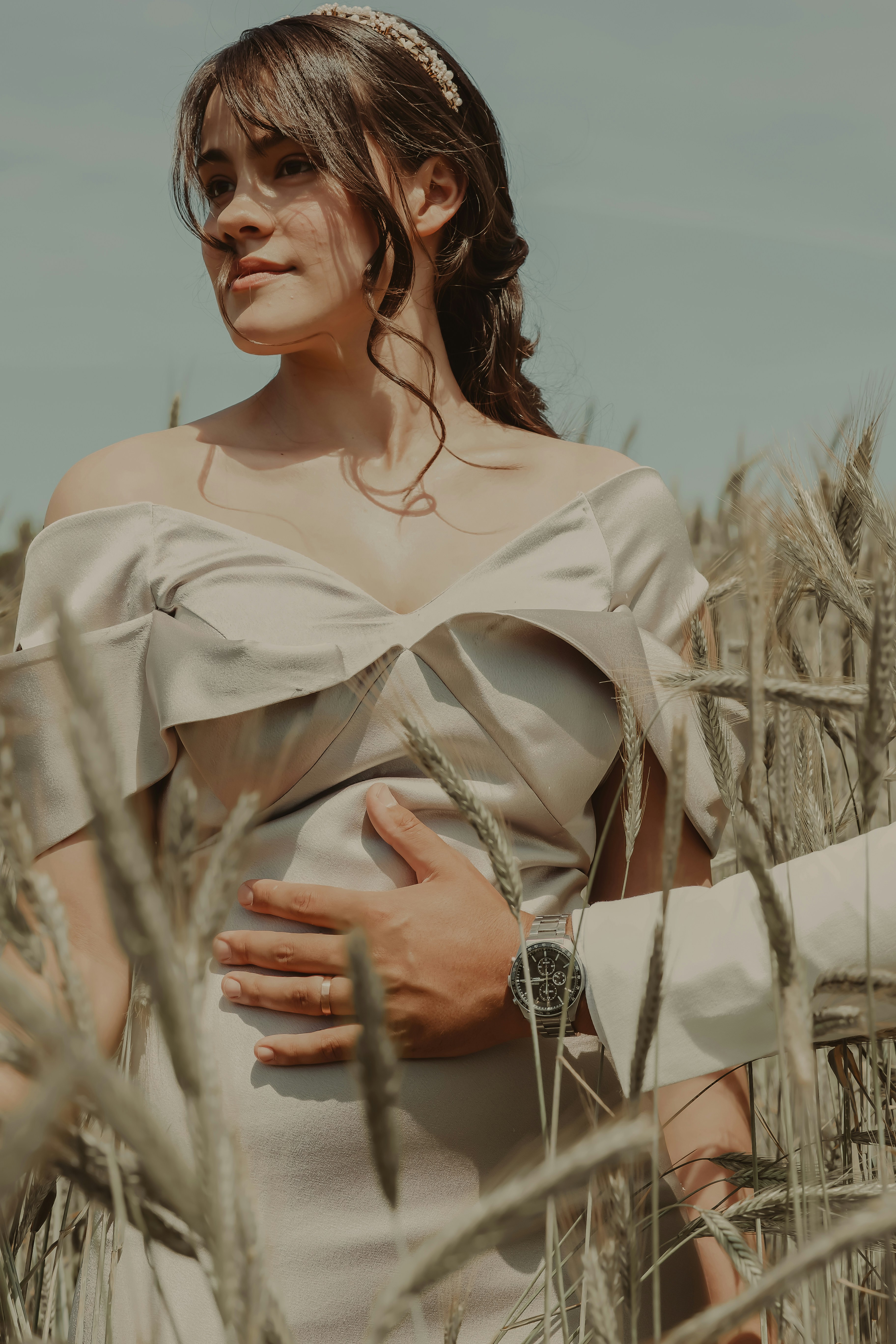 A couple stands in a field of wheat, with the woman in an elegant off-shoulder dress and the man gently holding her waist. The warm sunlight casts a soft glow on their intimate moment.