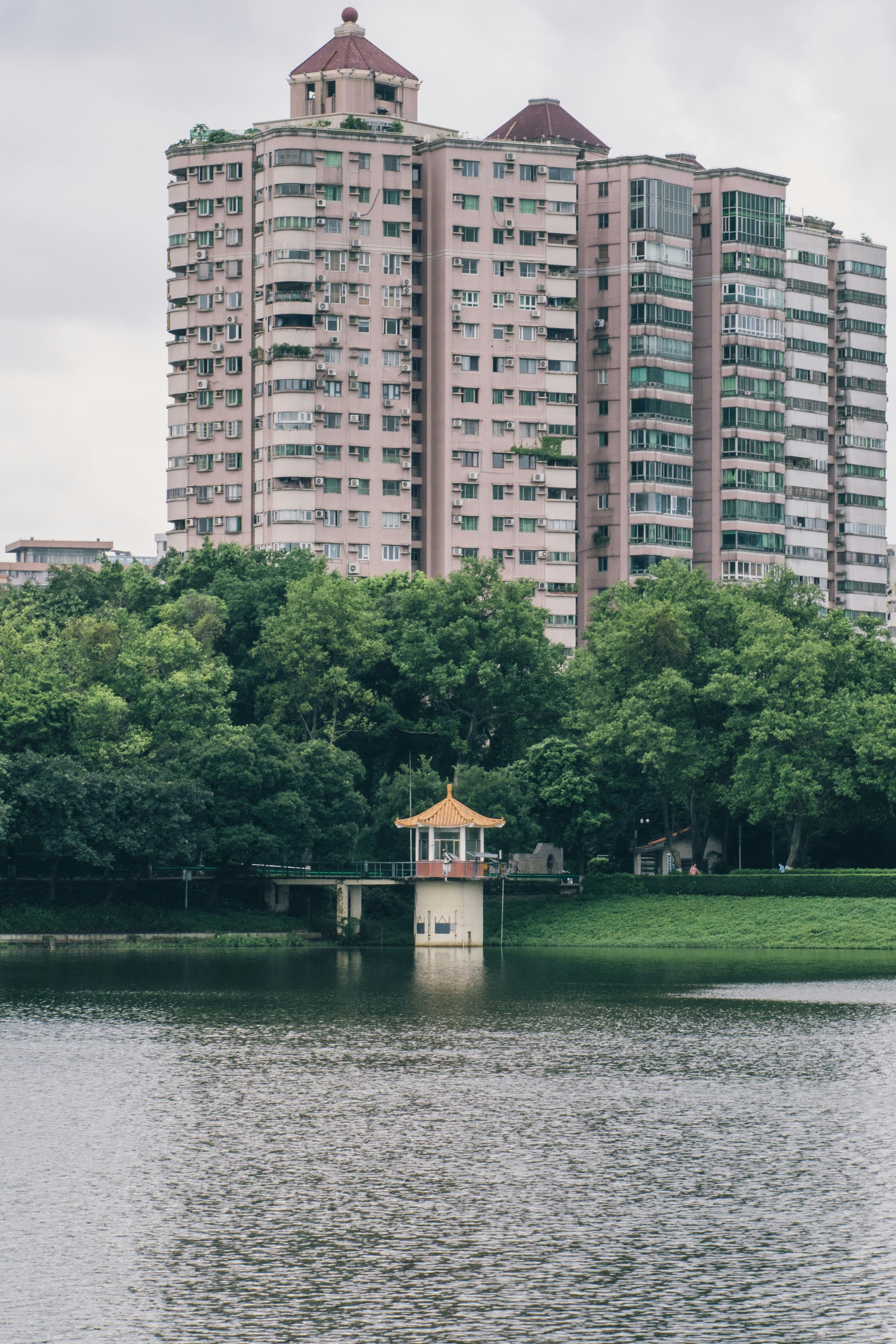 A serene lakeside pavilion nestled among lush greenery and towering residential buildings, reflecting a blend of urban life and natural beauty.