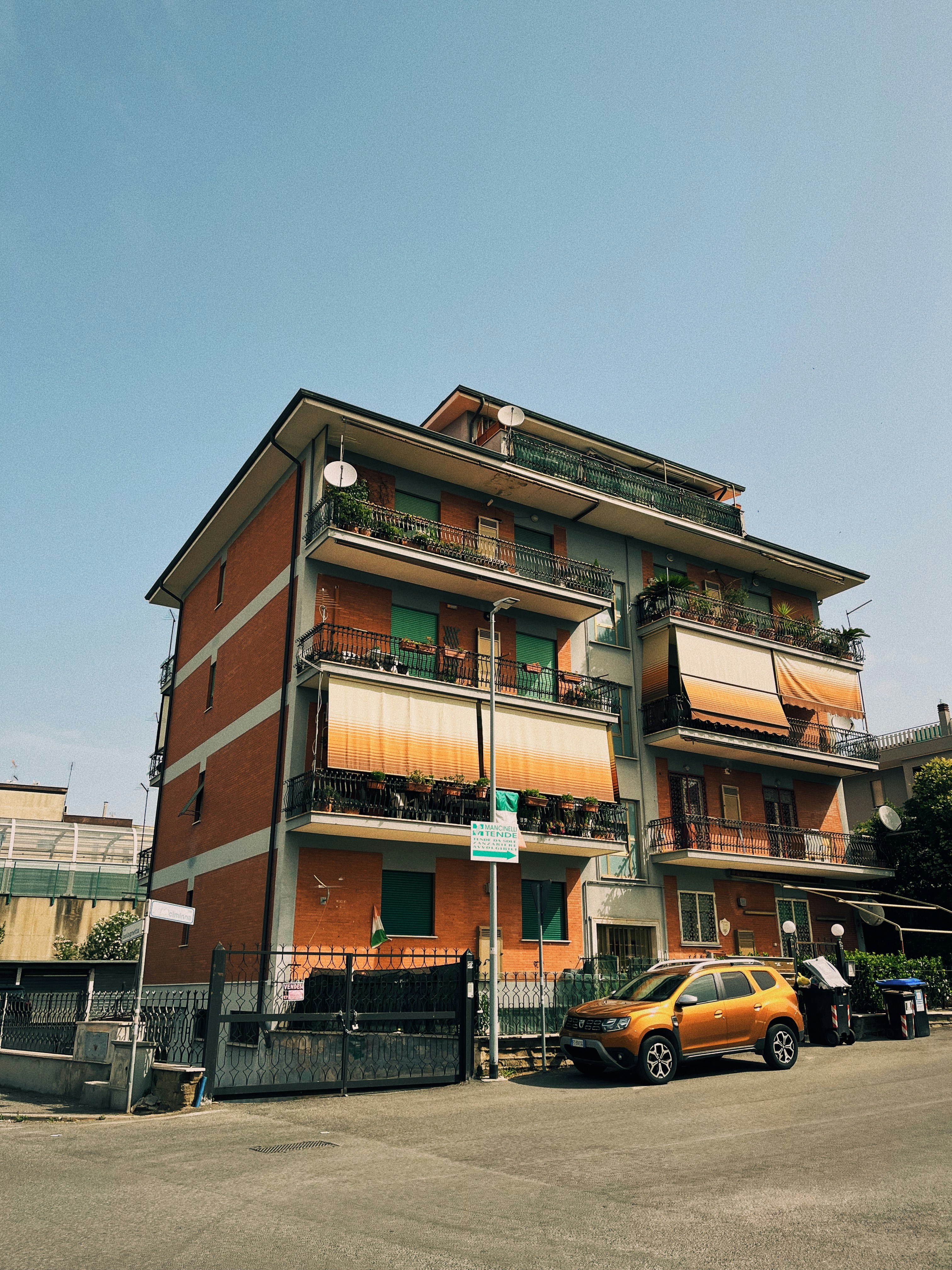 Colorful multi-story brick apartment building with balconies and orange awnings, parked car on the street, blue sky overhead.