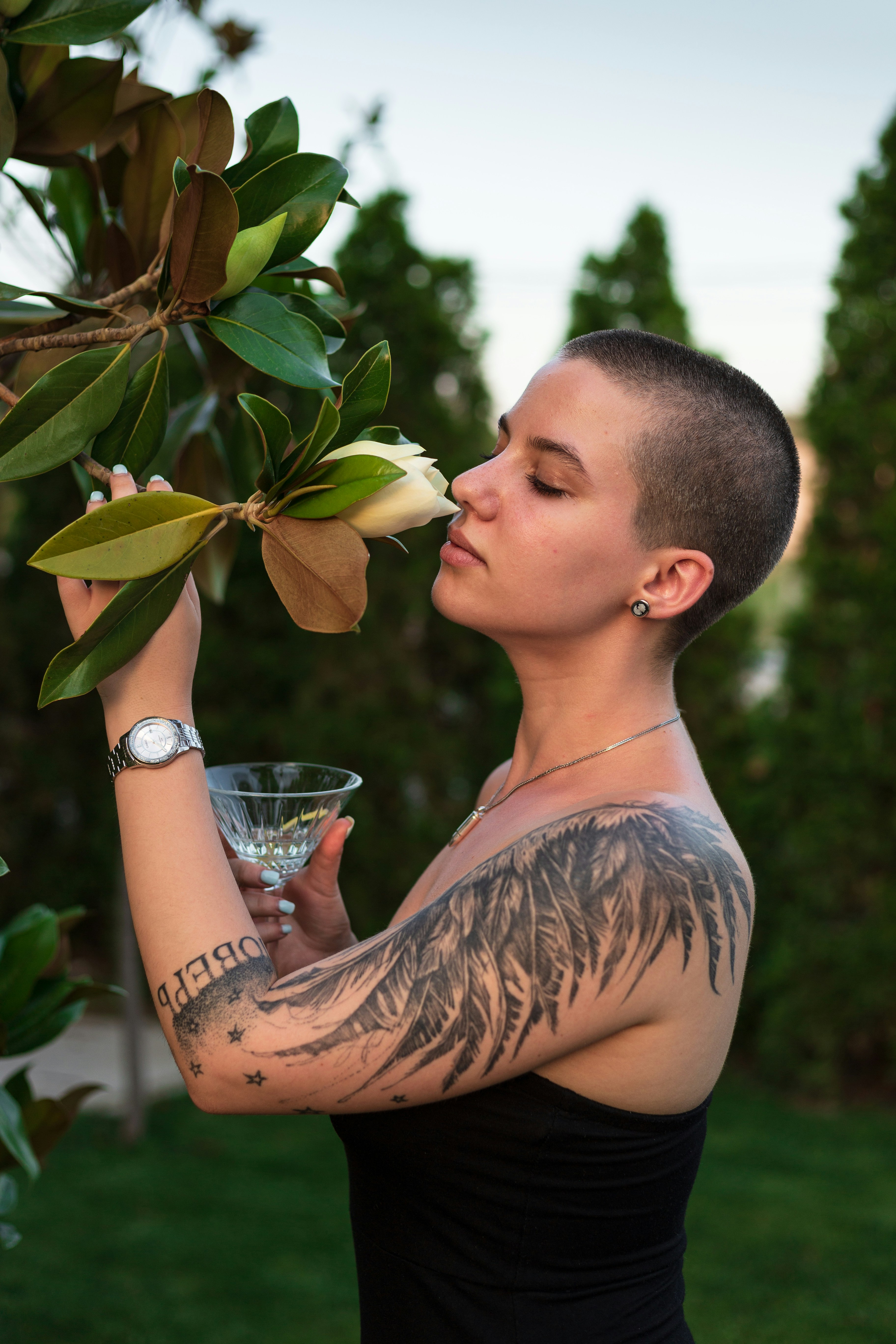 Woman with a tattoo gently touching a magnolia flower while holding a cocktail glass. The background features lush greenery.