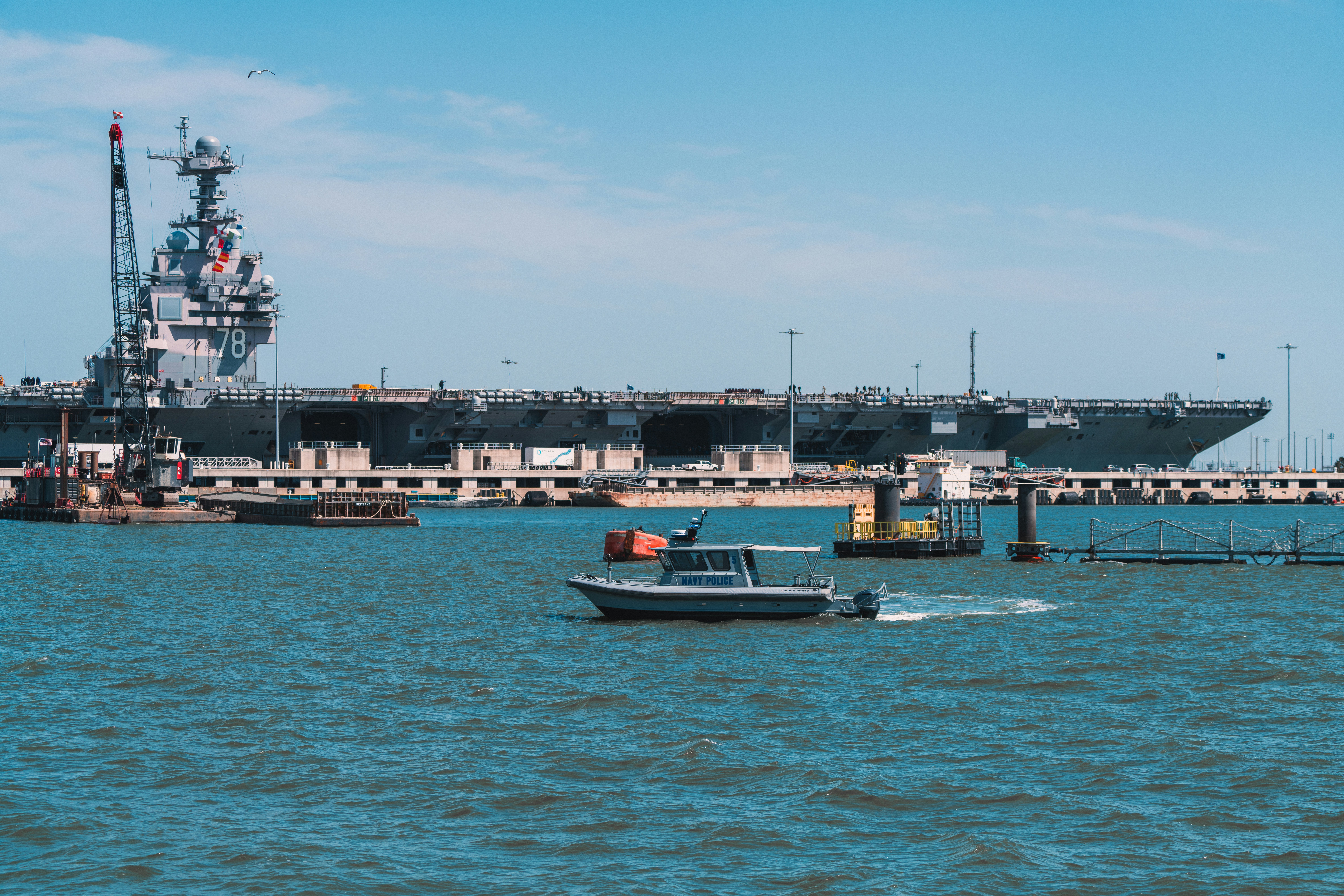 Navy patrol boat navigating near a massive aircraft carrier docked at a bustling harbor.