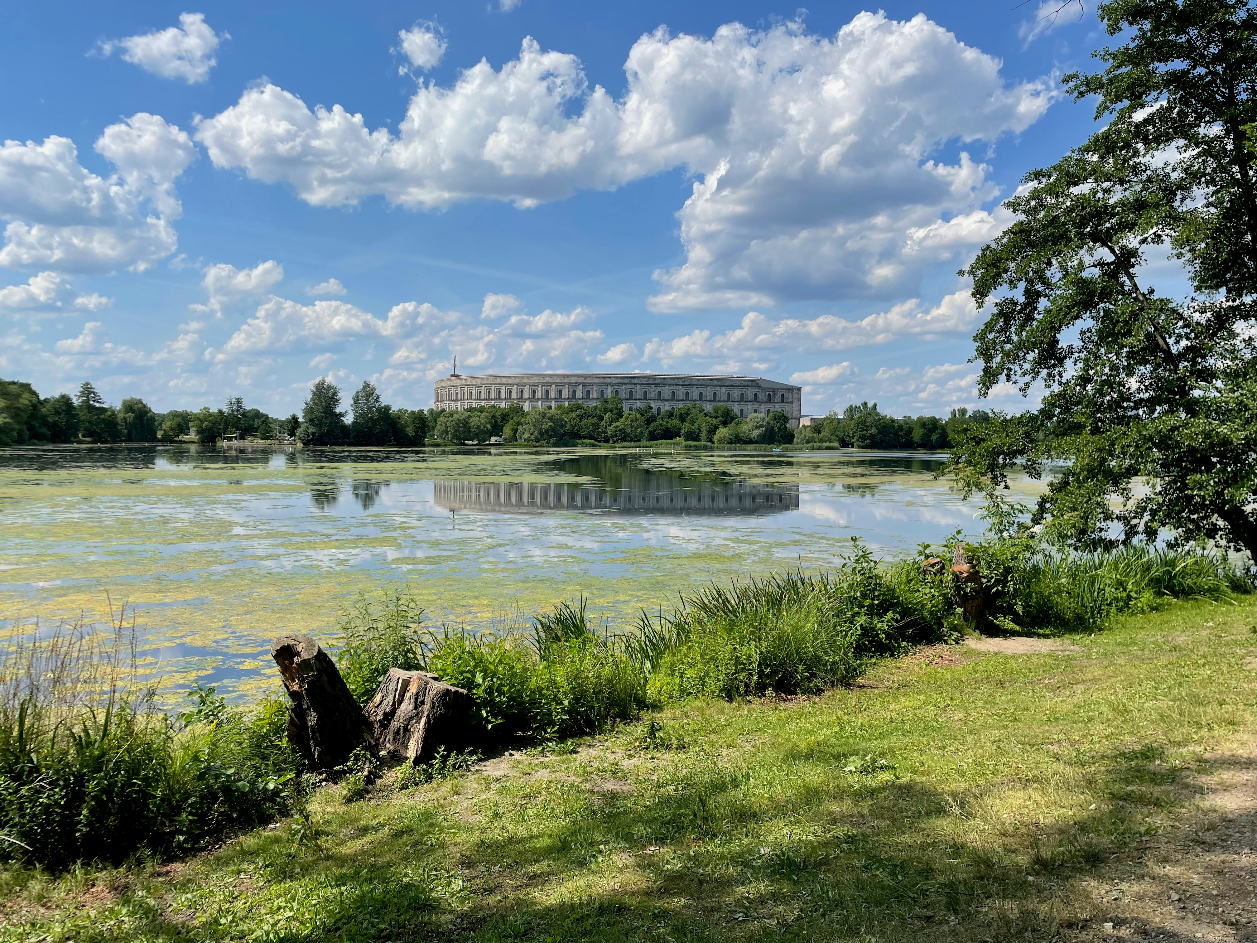 Tranquil lake reflecting a distant stadium under a bright blue sky with fluffy clouds.