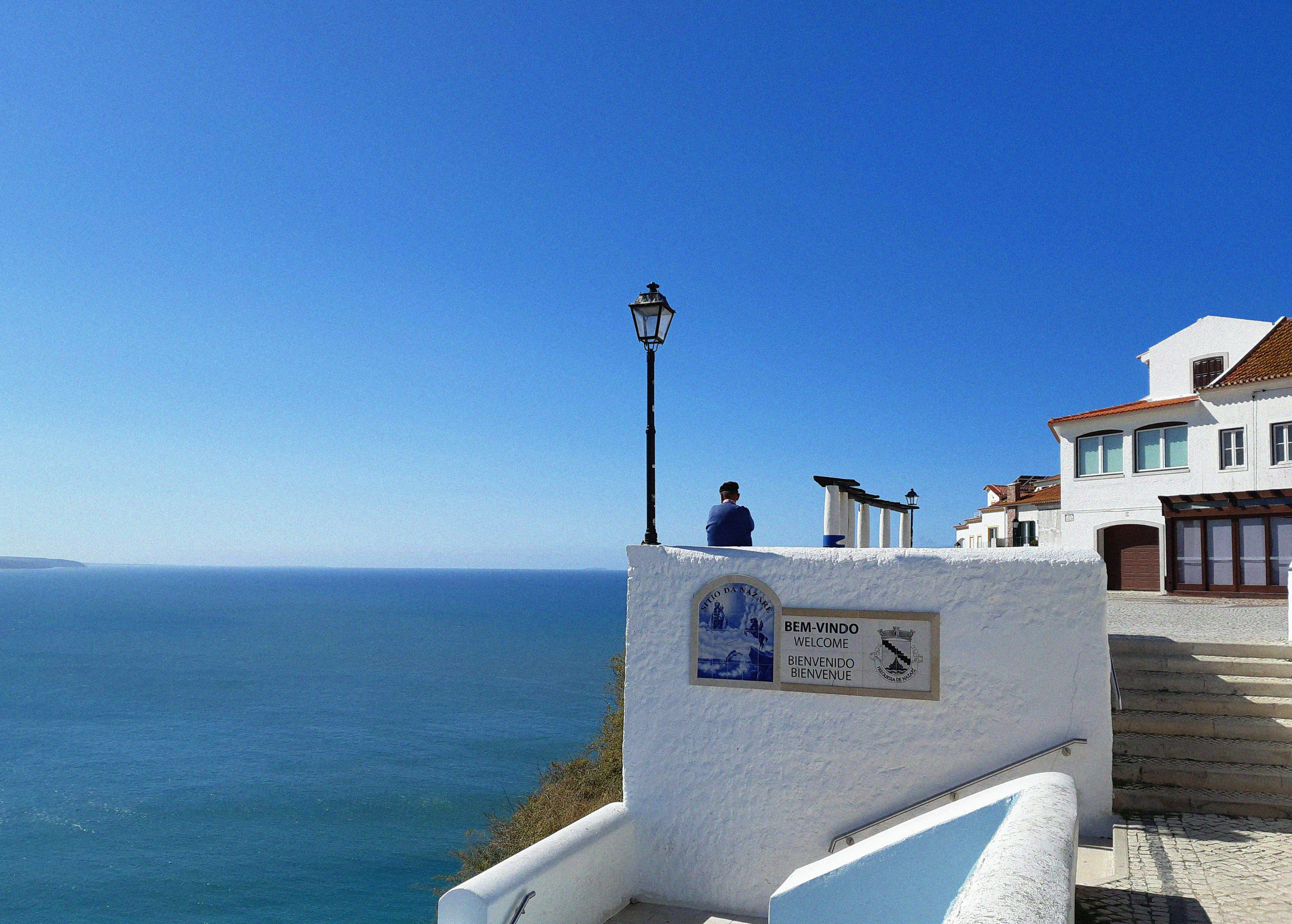 Whitewashed coastal terrace with steps and a railing extends toward a calm blue sea under a clear sky. A solitary figure stands near the wall, adding a sense of scale to the tranquil seascape.