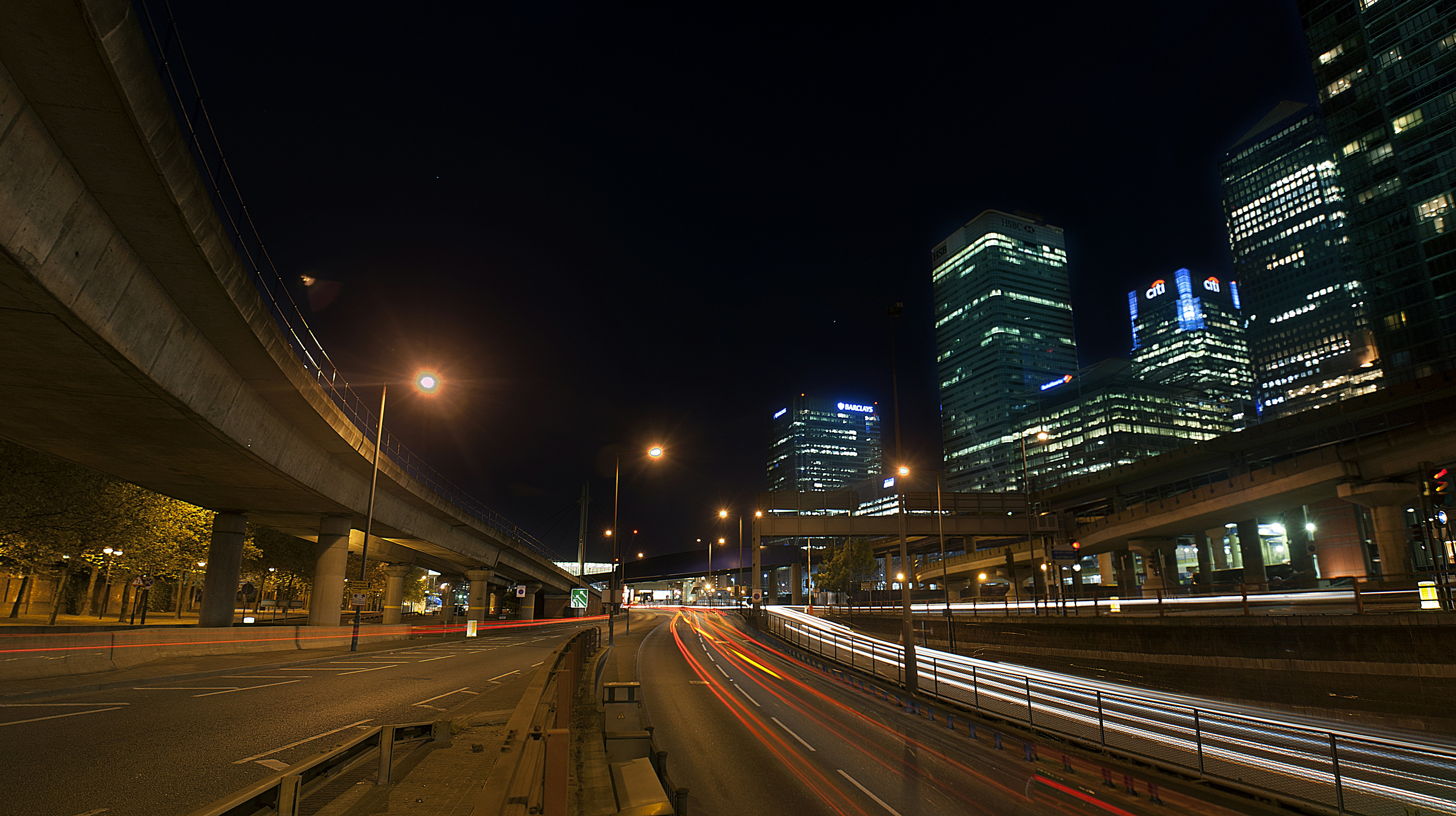 Long-exposure photograph of a nighttime cityscape with elevated highways and streaks of red and white lights. Modern glass towers rise in the background.