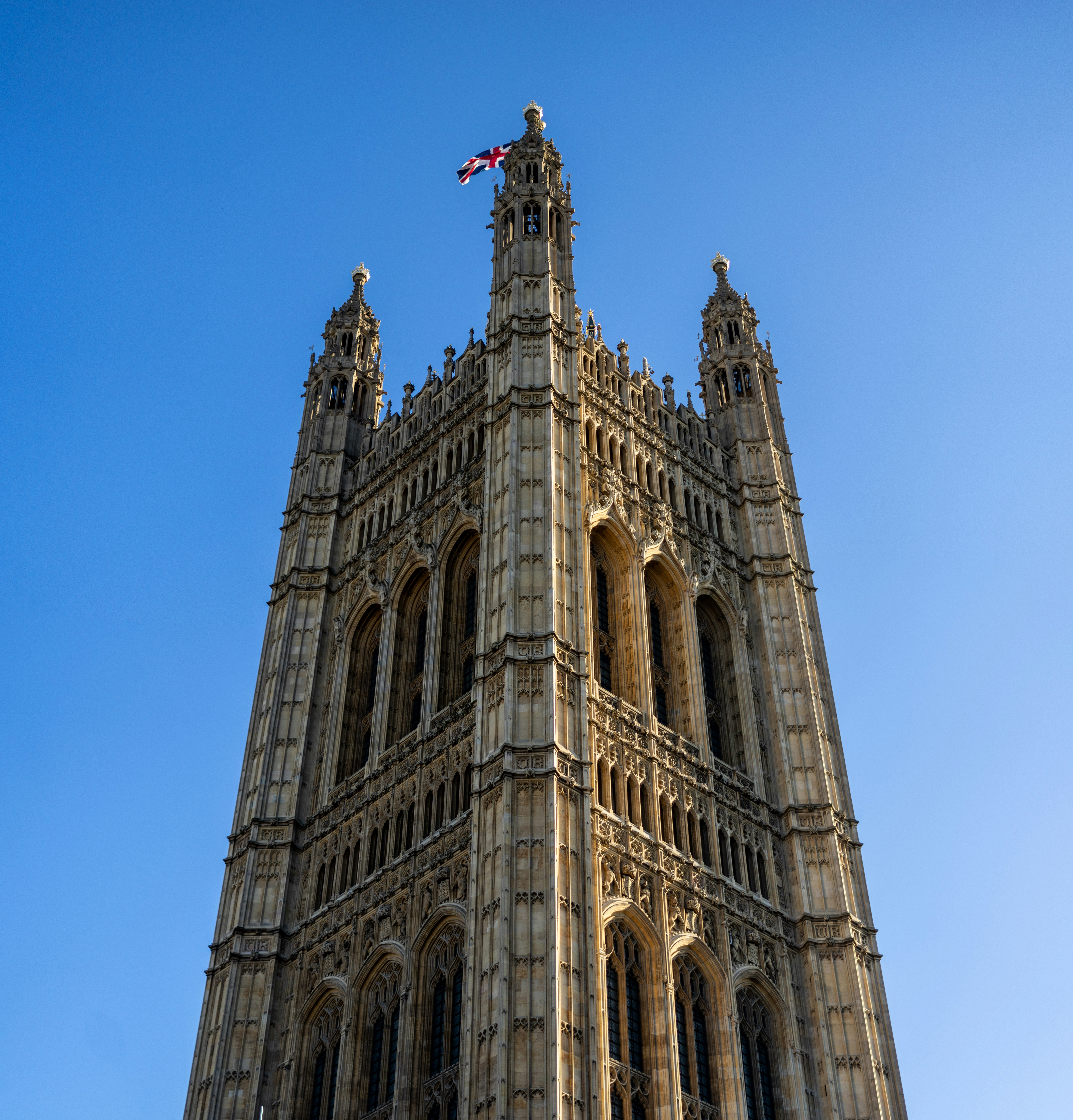 Gothic architecture of the Palace of Westminster's tower against a clear blue sky, showcasing intricate stonework and a fluttering flag.