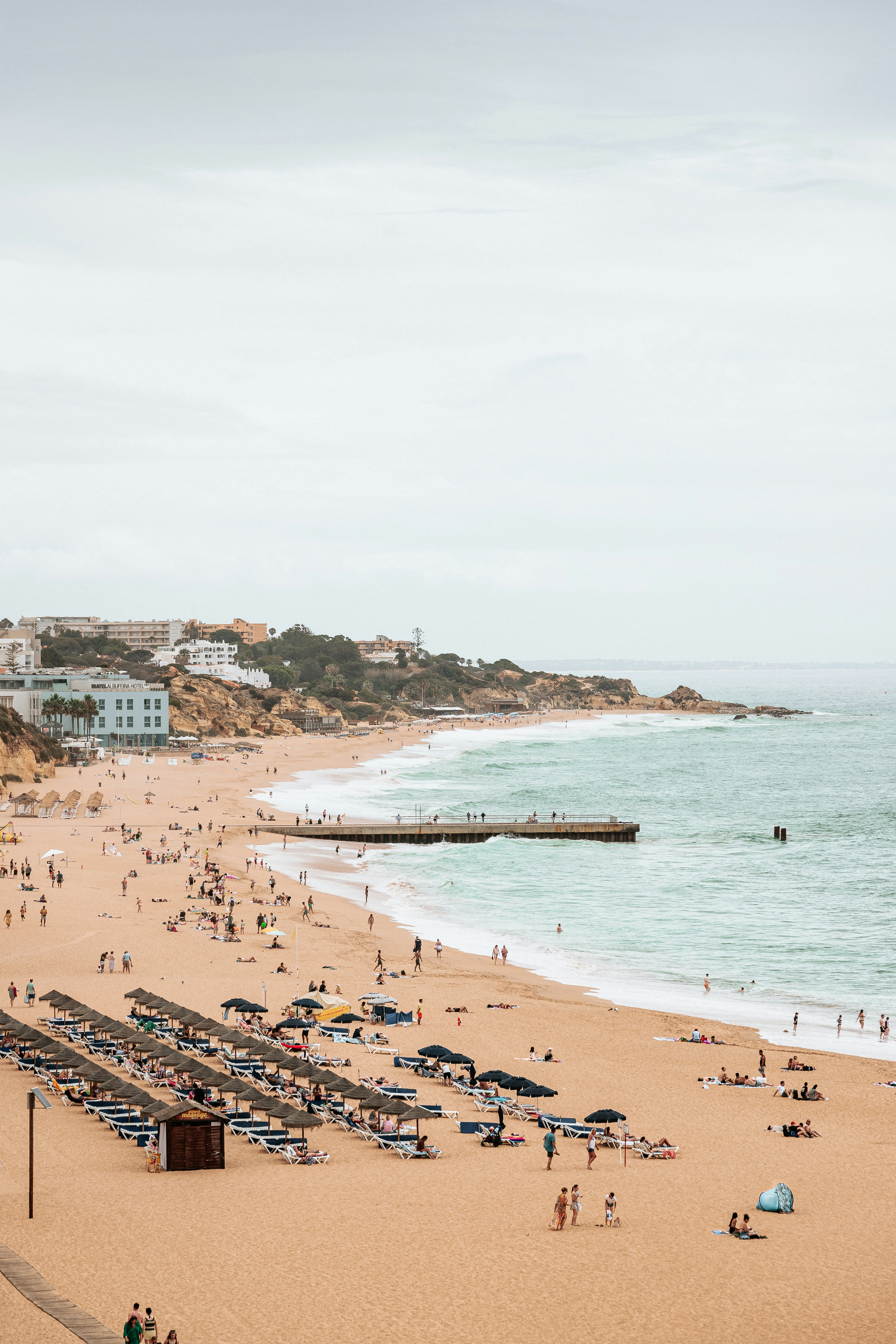 Crowded beach with sunbathers and umbrellas along the sandy coast of Albufeira under a cloudy sky.