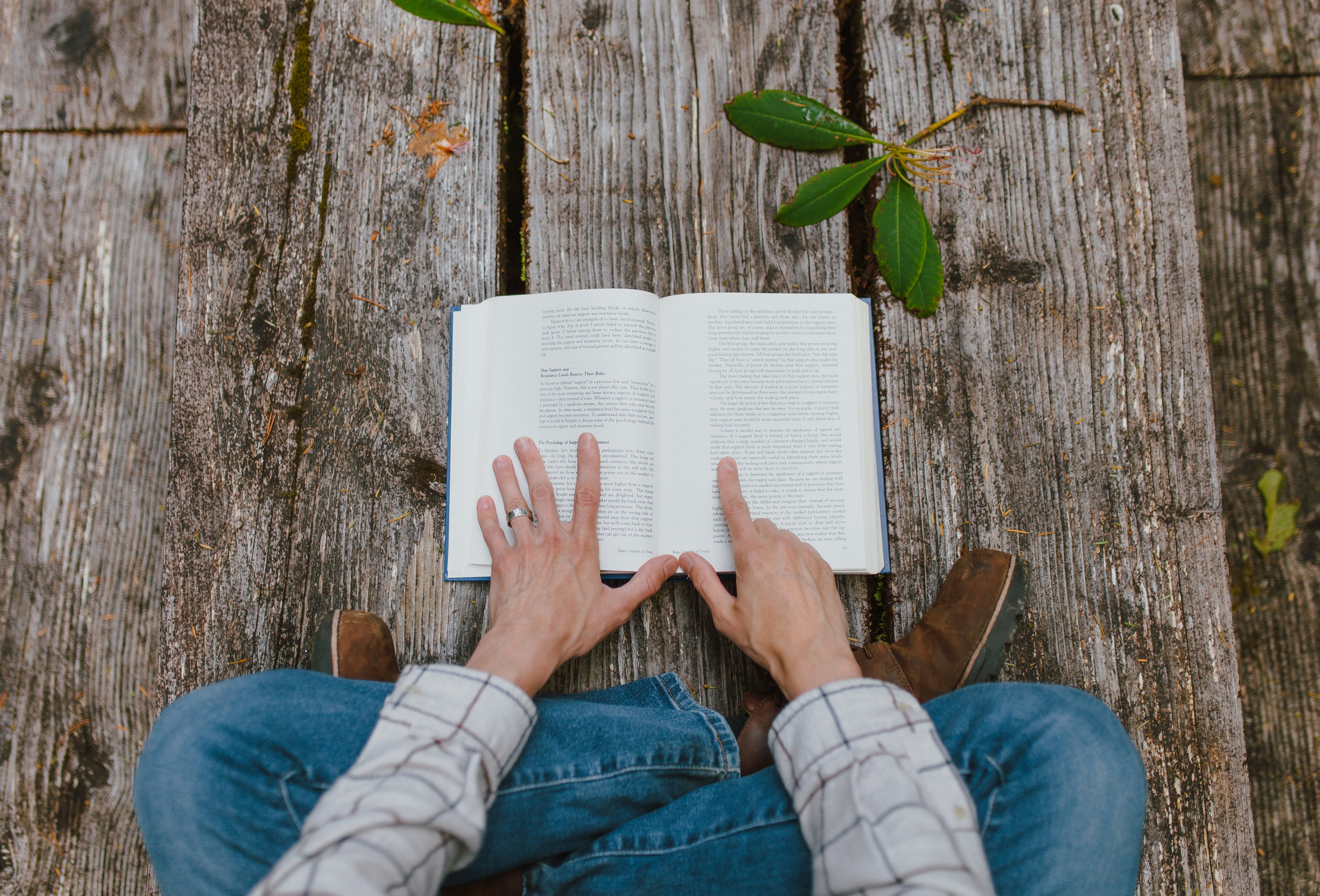 Open book resting on a rustic wooden surface, with hands gently pointing at the text. Green leaves add a touch of nature.
