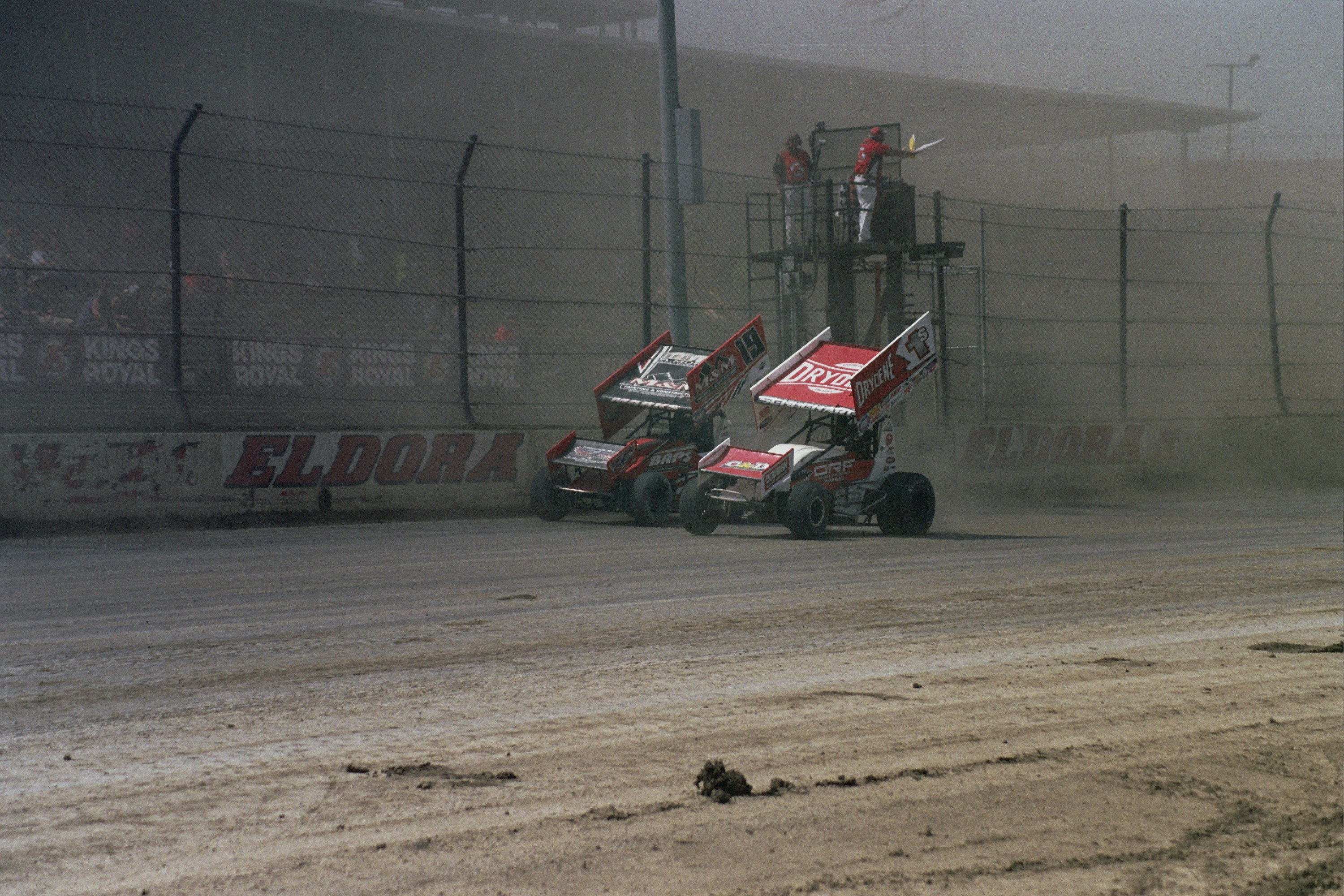 Two sprint cars racing side-by-side on a dirt track, kicking up clouds of dust.
