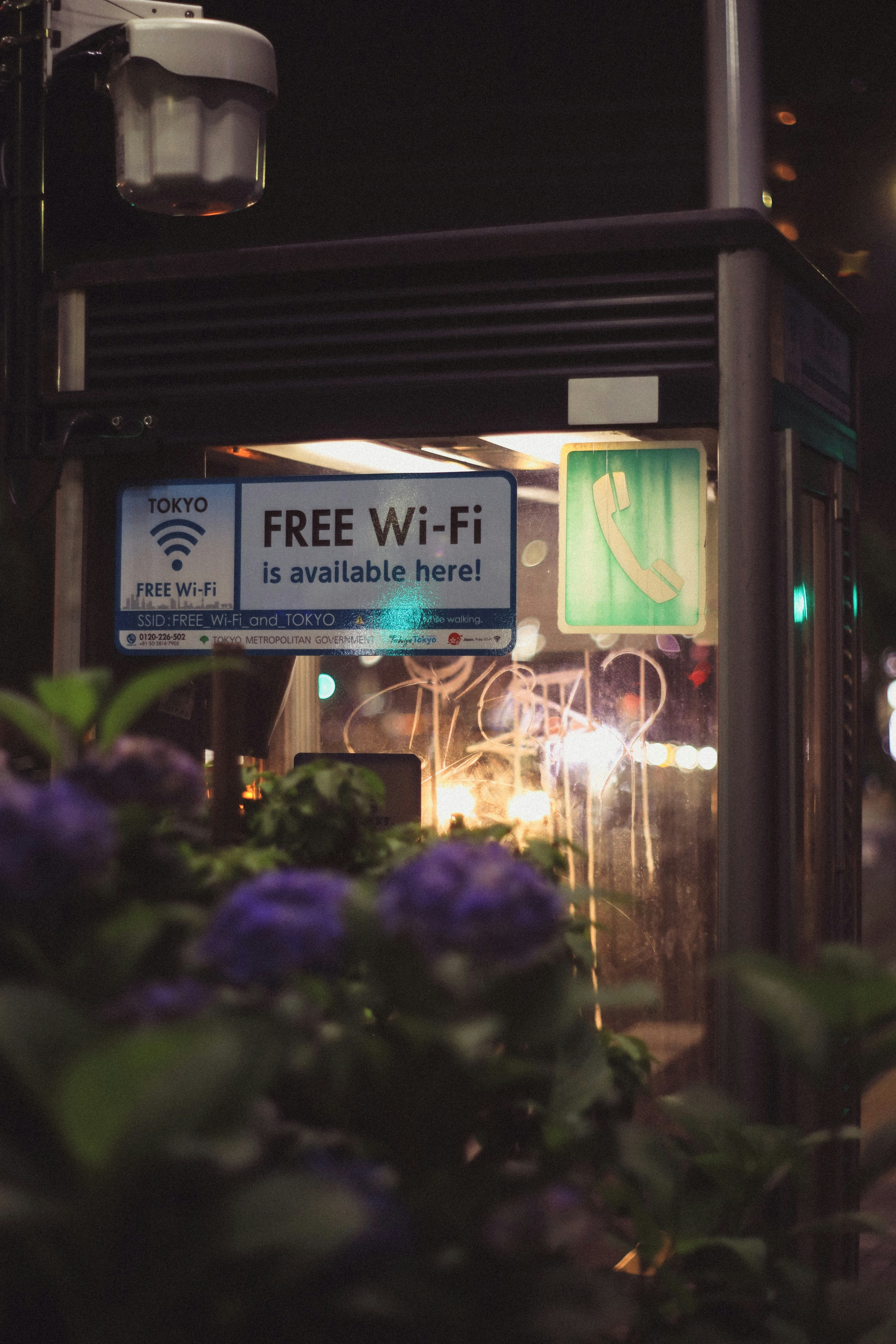 A phone booth in Tokyo featuring a sign that advertises free Wi-Fi, surrounded by lush greenery. The scene captures the blend of technology and nature in a bustling city environment.