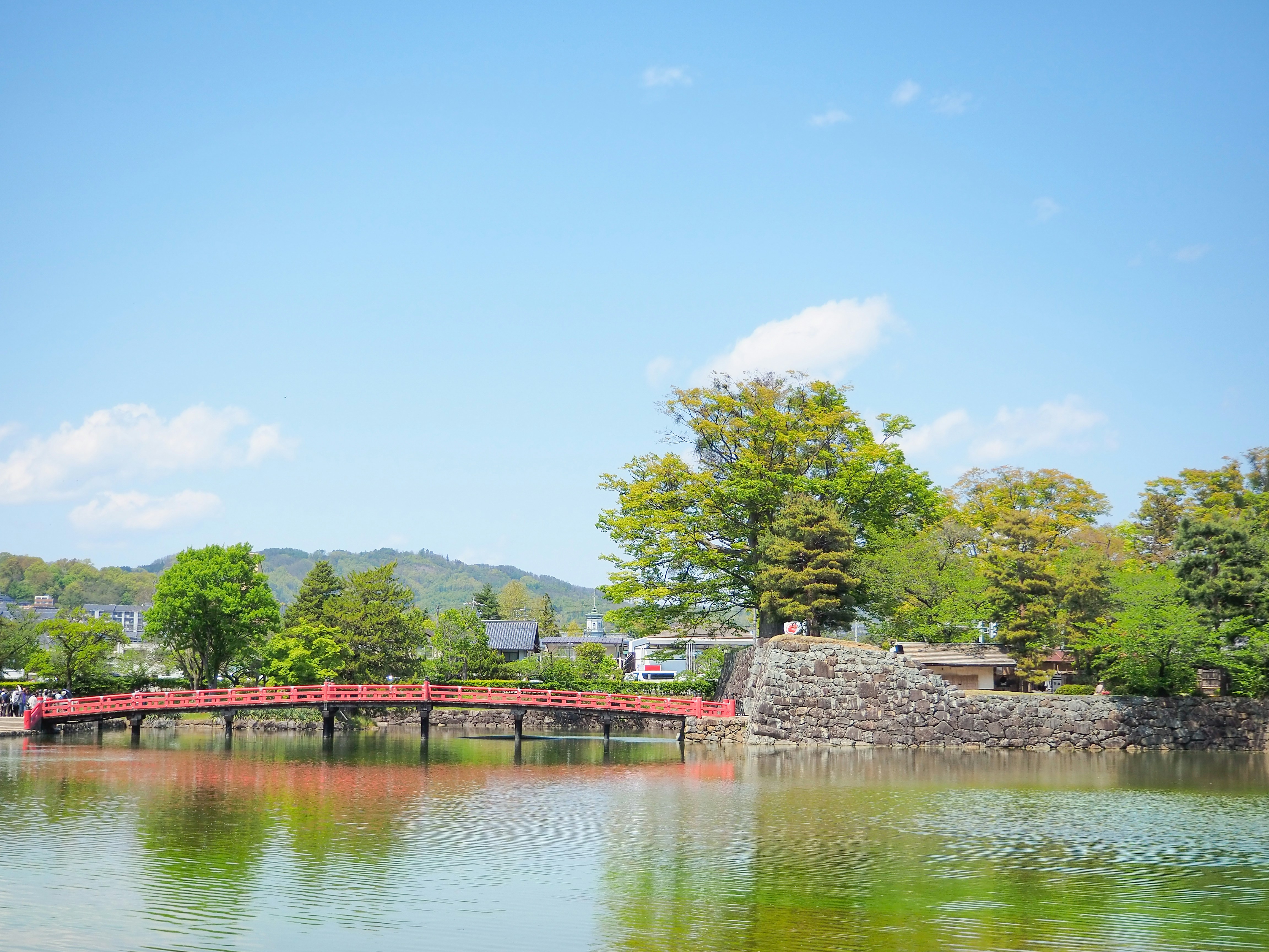 A vibrant red bridge arches over a tranquil pond, surrounded by lush greenery and a stone wall, under a clear blue sky.
