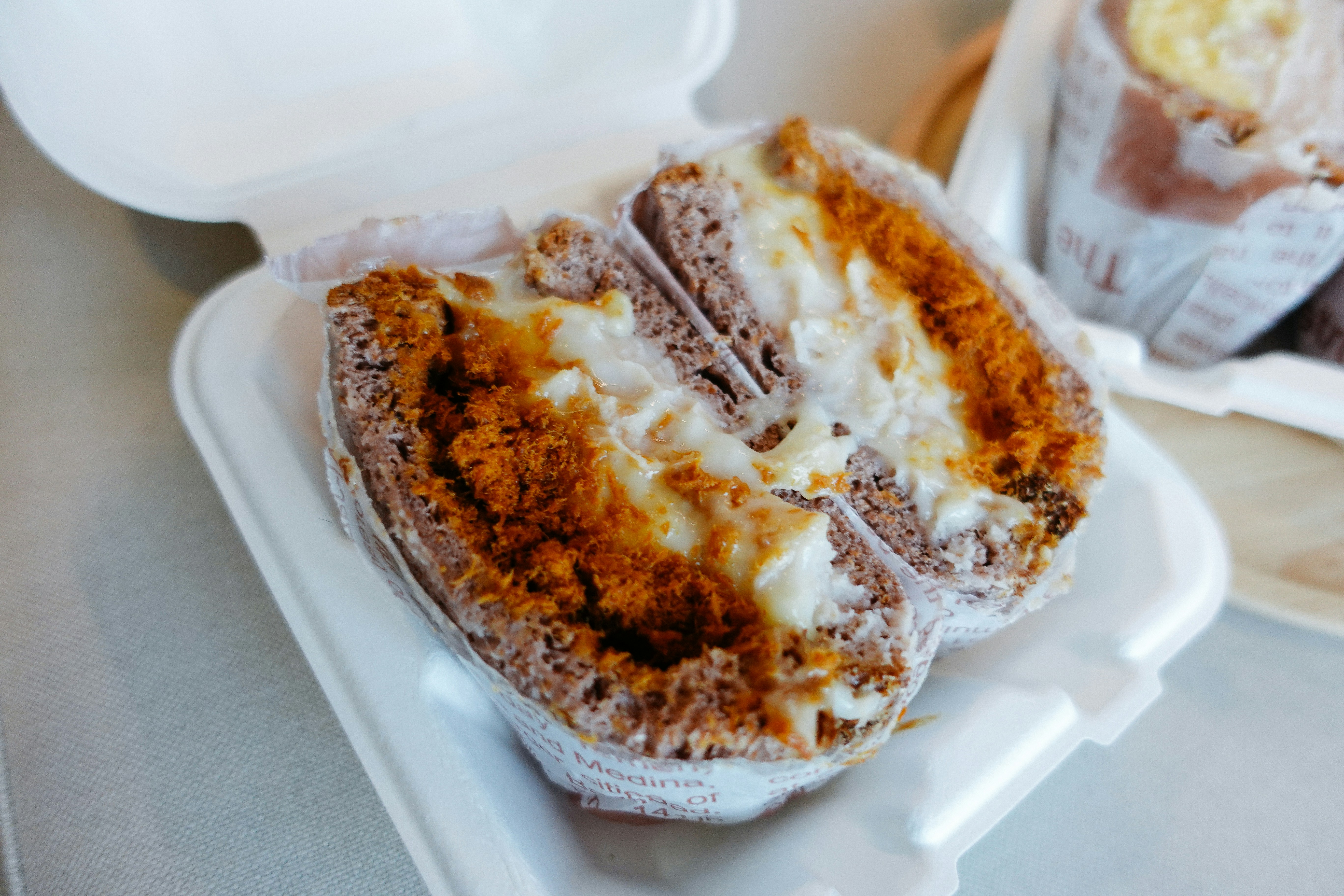 A split pastry sits in a white takeout box with visible creamy filling and cinnamon glaze, ready for a snack. This is food photography of a baked goods item.