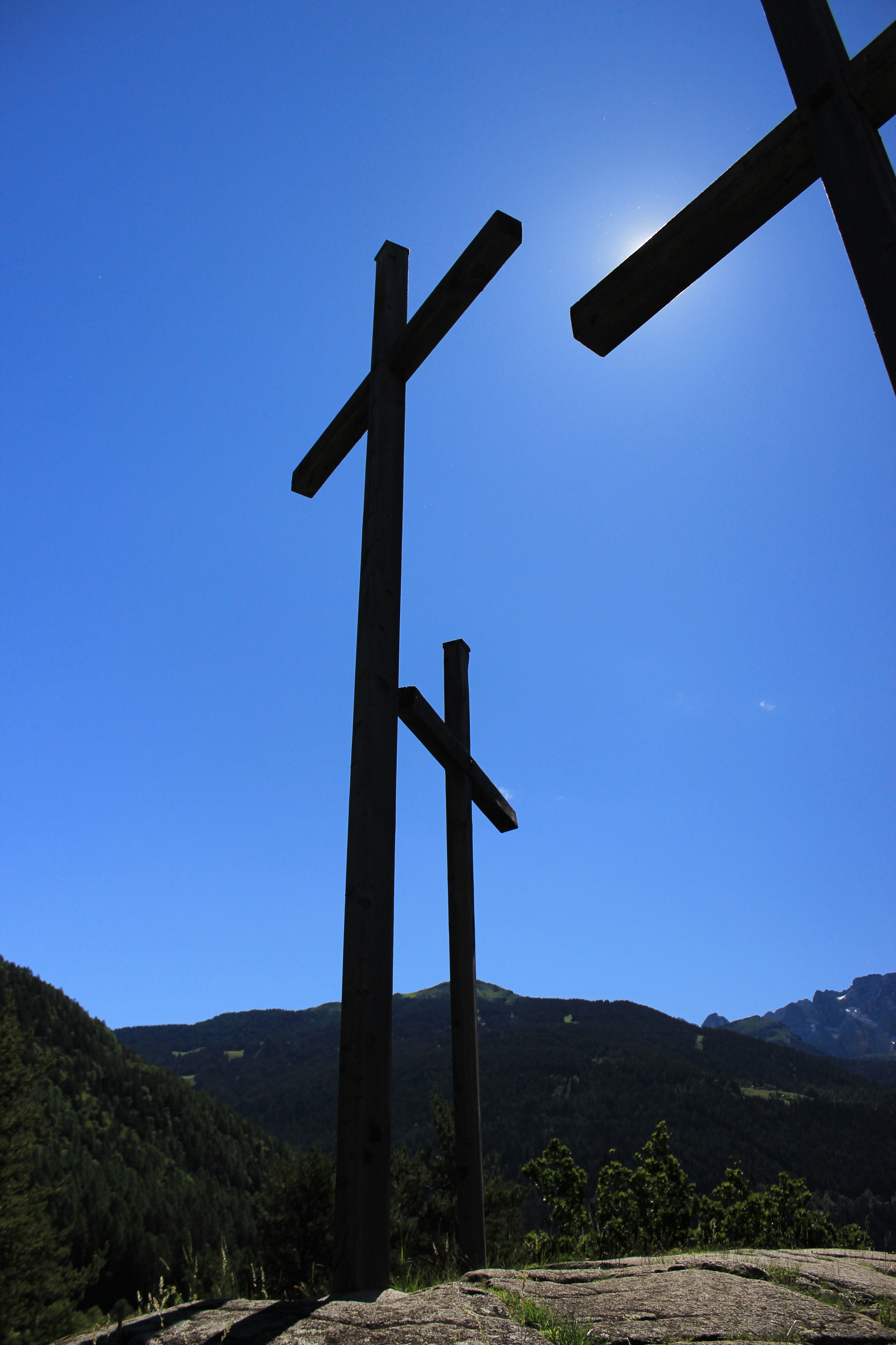 Three towering wooden crosses silhouetted against a clear blue sky, set against a backdrop of mountains and lush greenery.