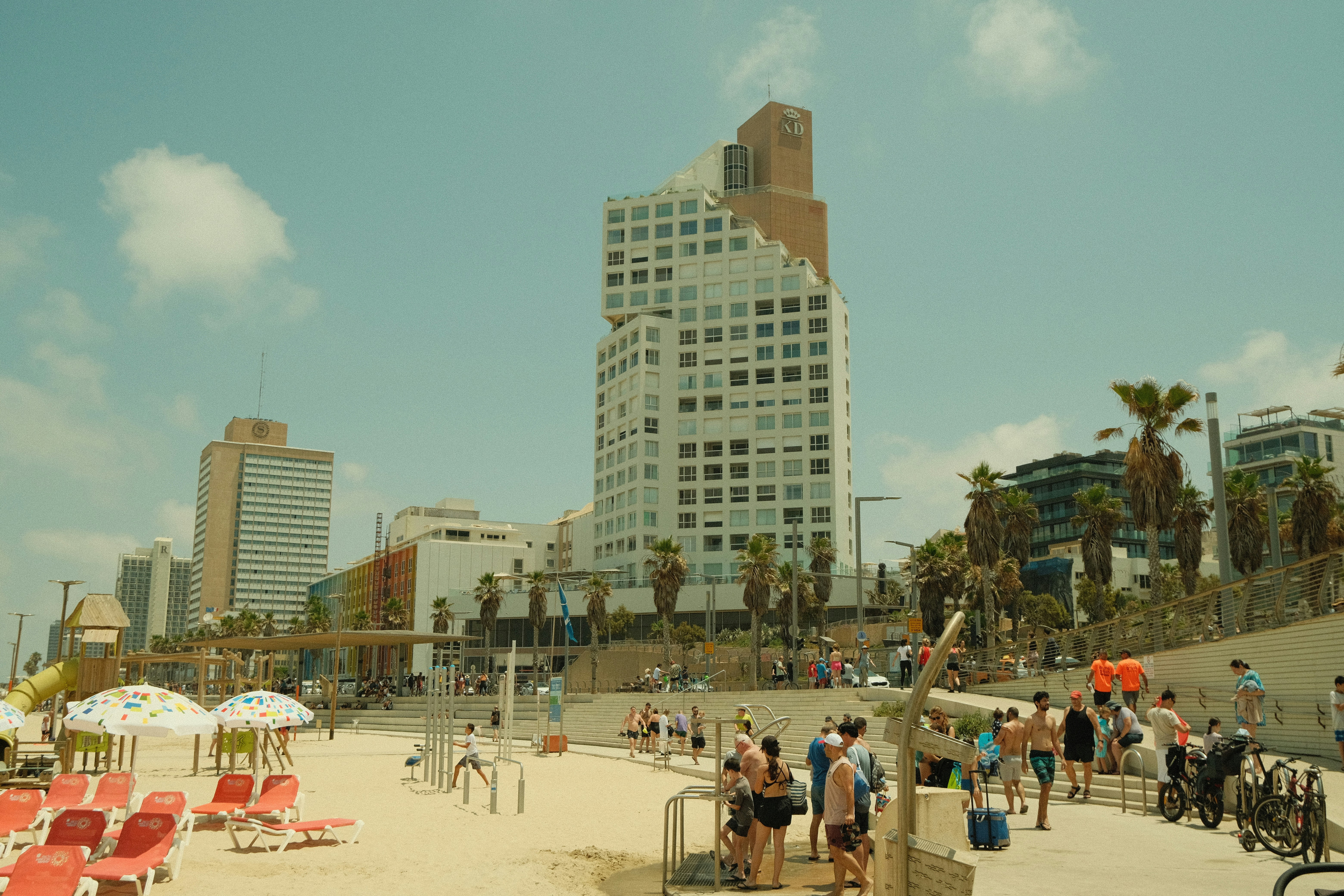Beachgoers enjoy a sunny day near modern high-rise buildings and palm trees.