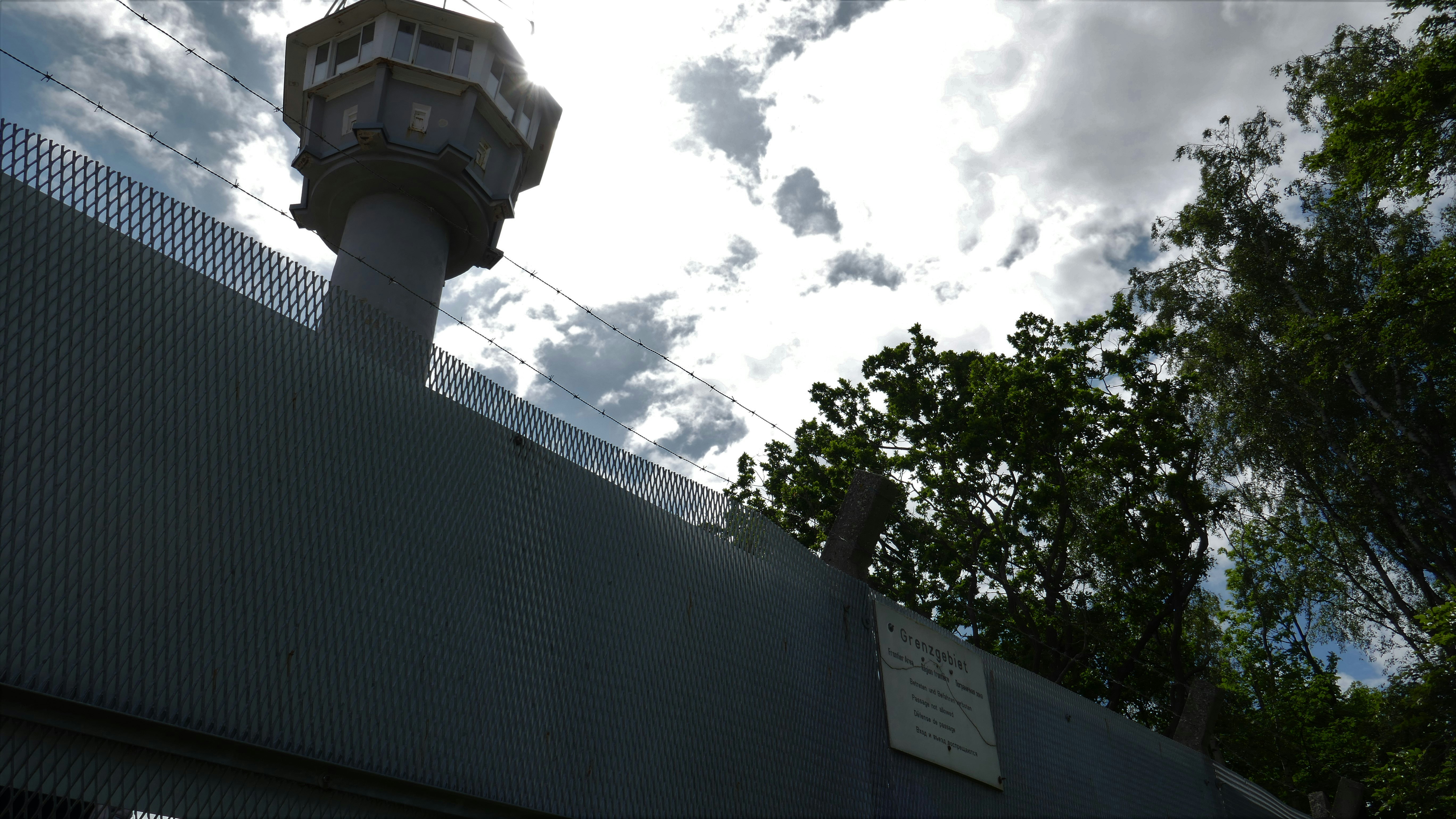 A watchtower looms over a security fence, framed by a dynamic sky filled with clouds. The scene conveys a sense of vigilance and protection.