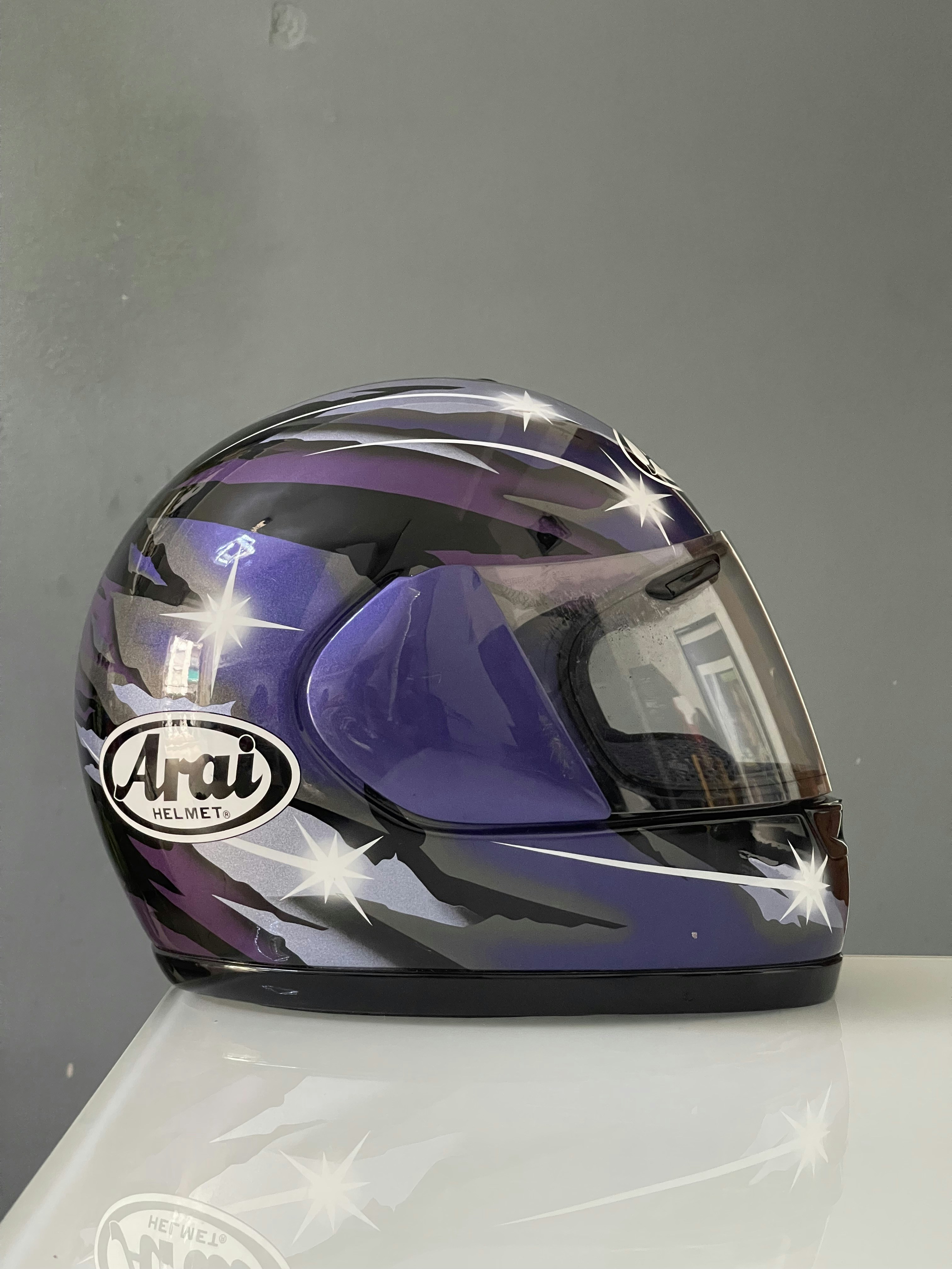 A purple and black Arai helmet with striking designs sits on a reflective surface against a gray backdrop.