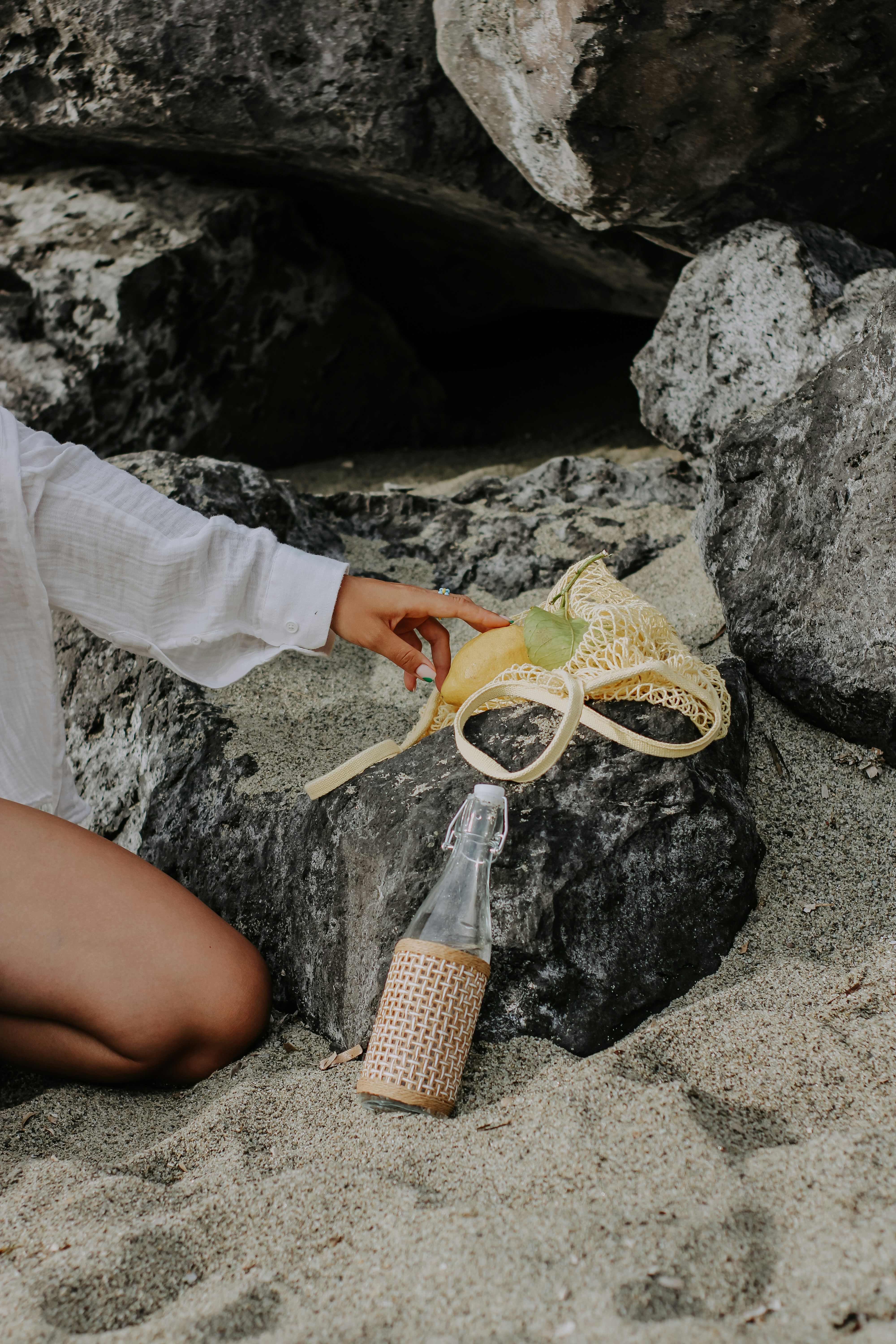 Hand reaches for a pale lemon and straw hat resting beside a glass bottled drink on a sandy beach, with rough rocks in the background. A muted, natural scene with warm tones.