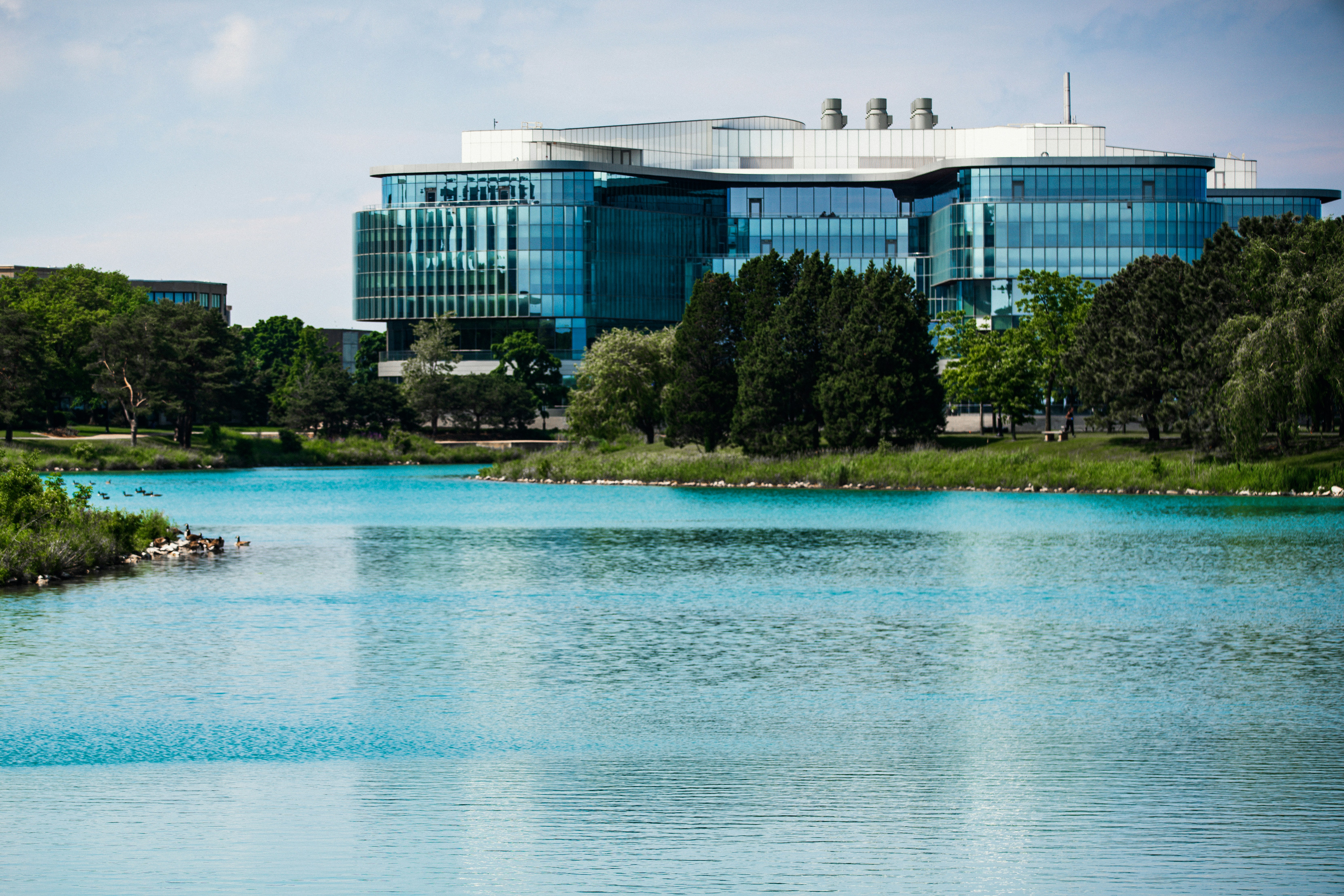 Modern glass building reflecting in a serene blue lake surrounded by lush trees.