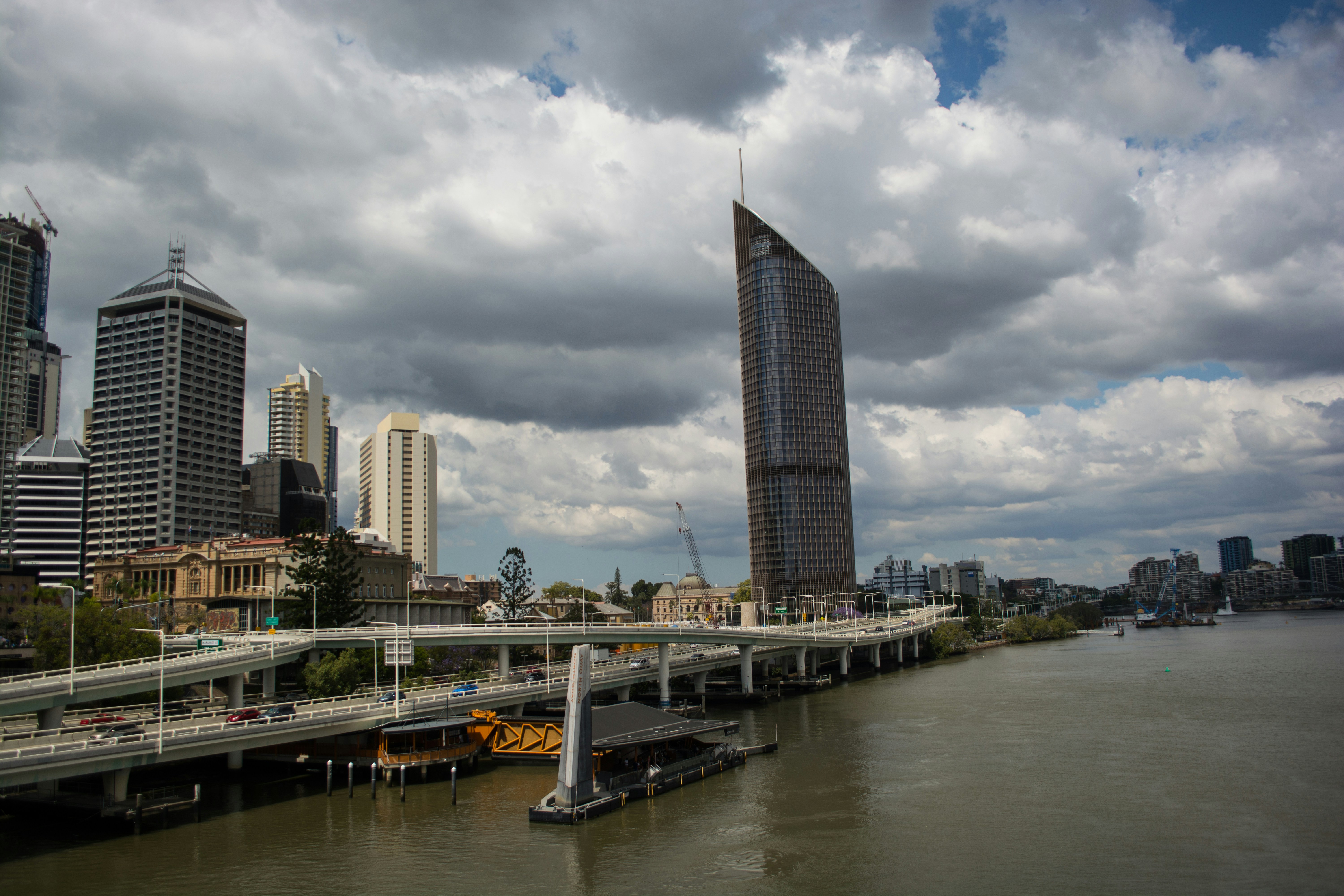 A contemporary skyline with a mix of modern and historical architecture alongside a river, under a dramatic cloudscape. The scene captures the essence of urban life intertwined with natural elements.