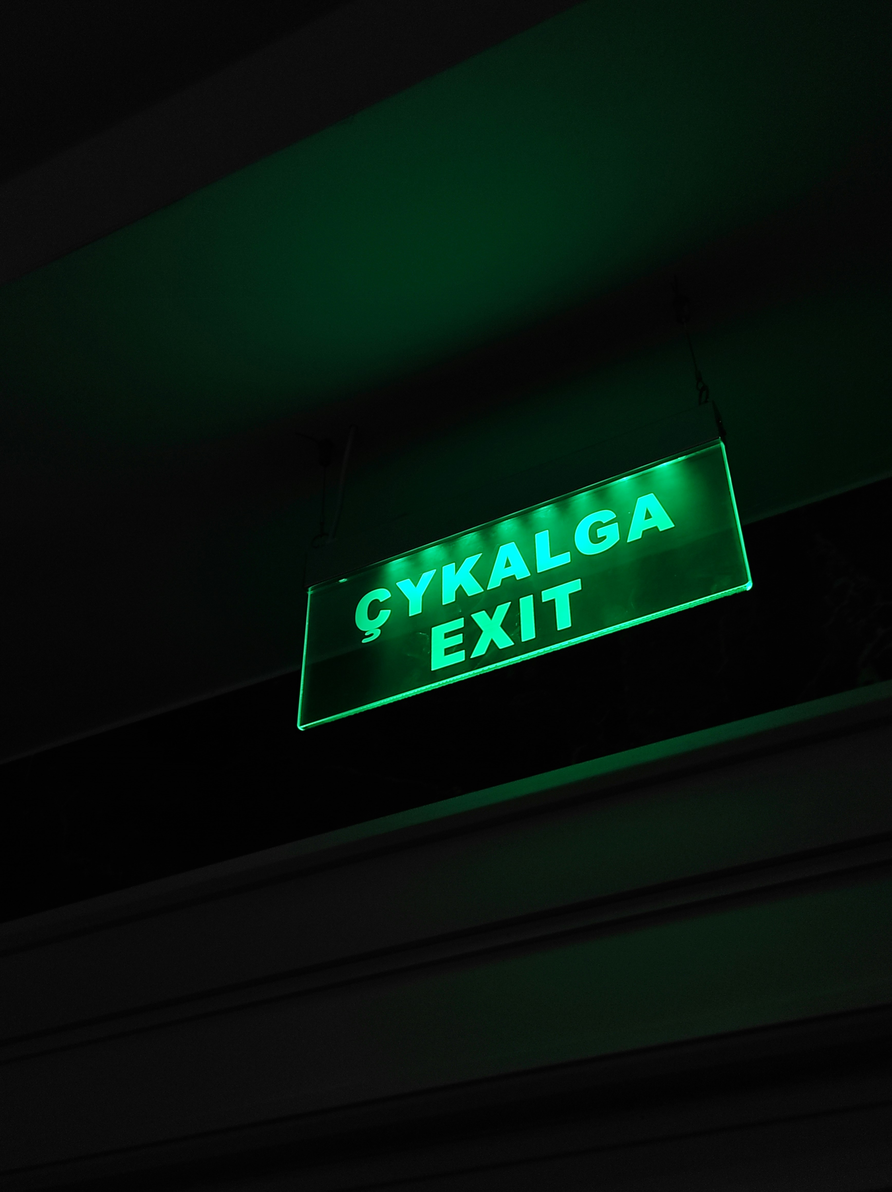 Green exit sign with the word 'ÇYKALGA' illuminated, hanging against a dark background.