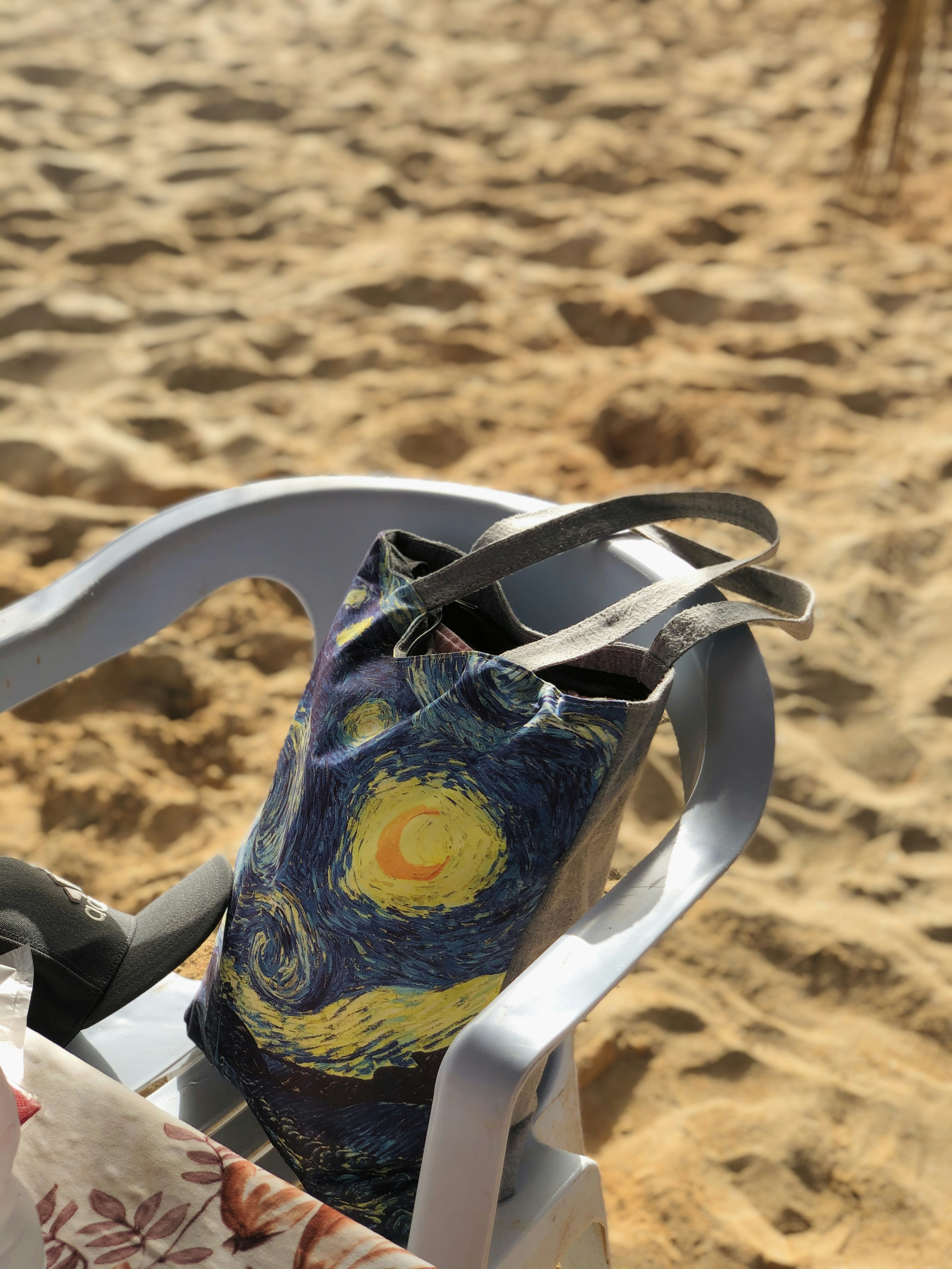A beach bag featuring Van Gogh's Starry Night rests on a plastic chair, surrounded by soft sand. The scene captures a moment of leisure and artistic flair.