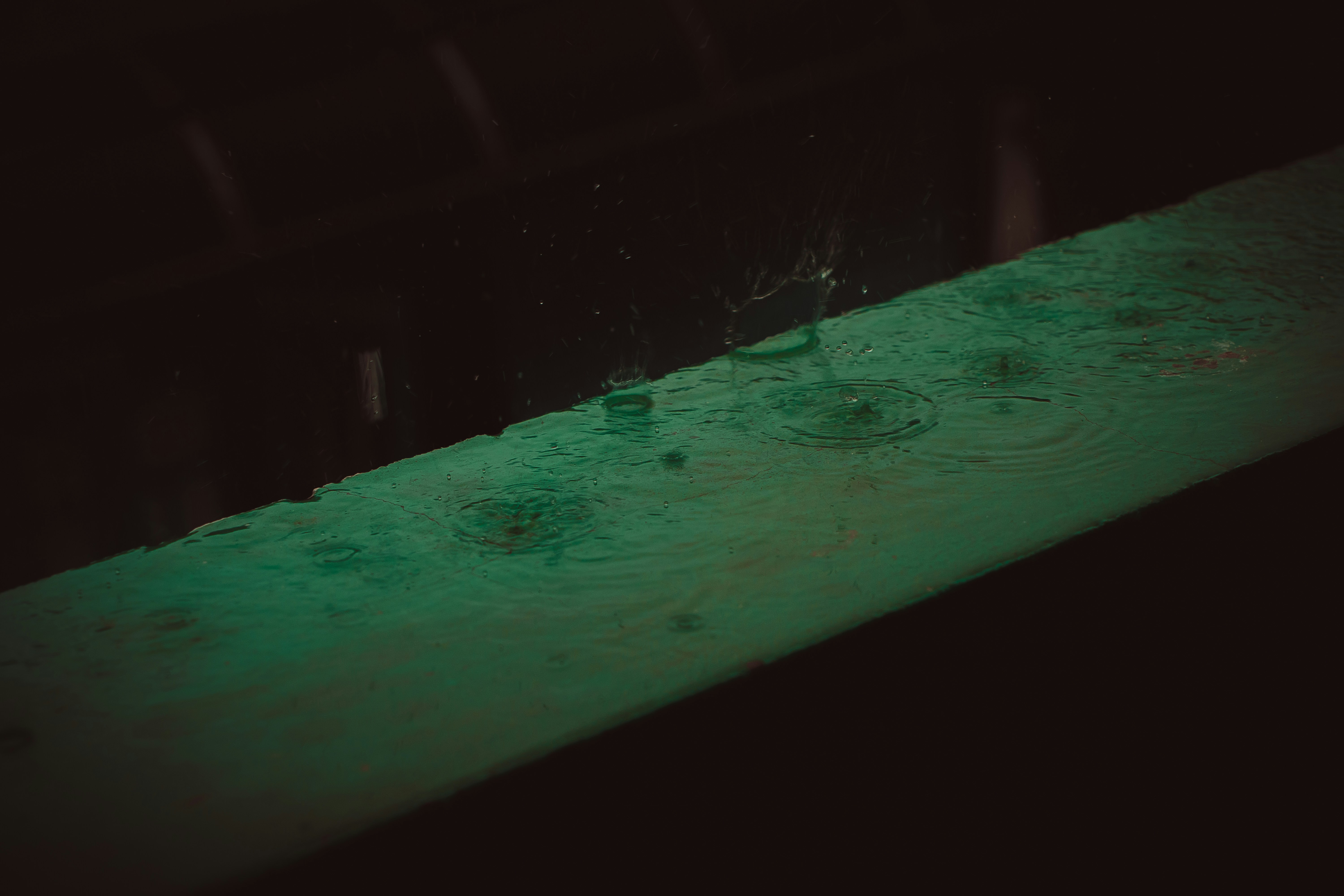 Raindrops create concentric ripples on a green-tinted surface, illuminated by soft light. The dark background enhances the vibrant colors and textures.