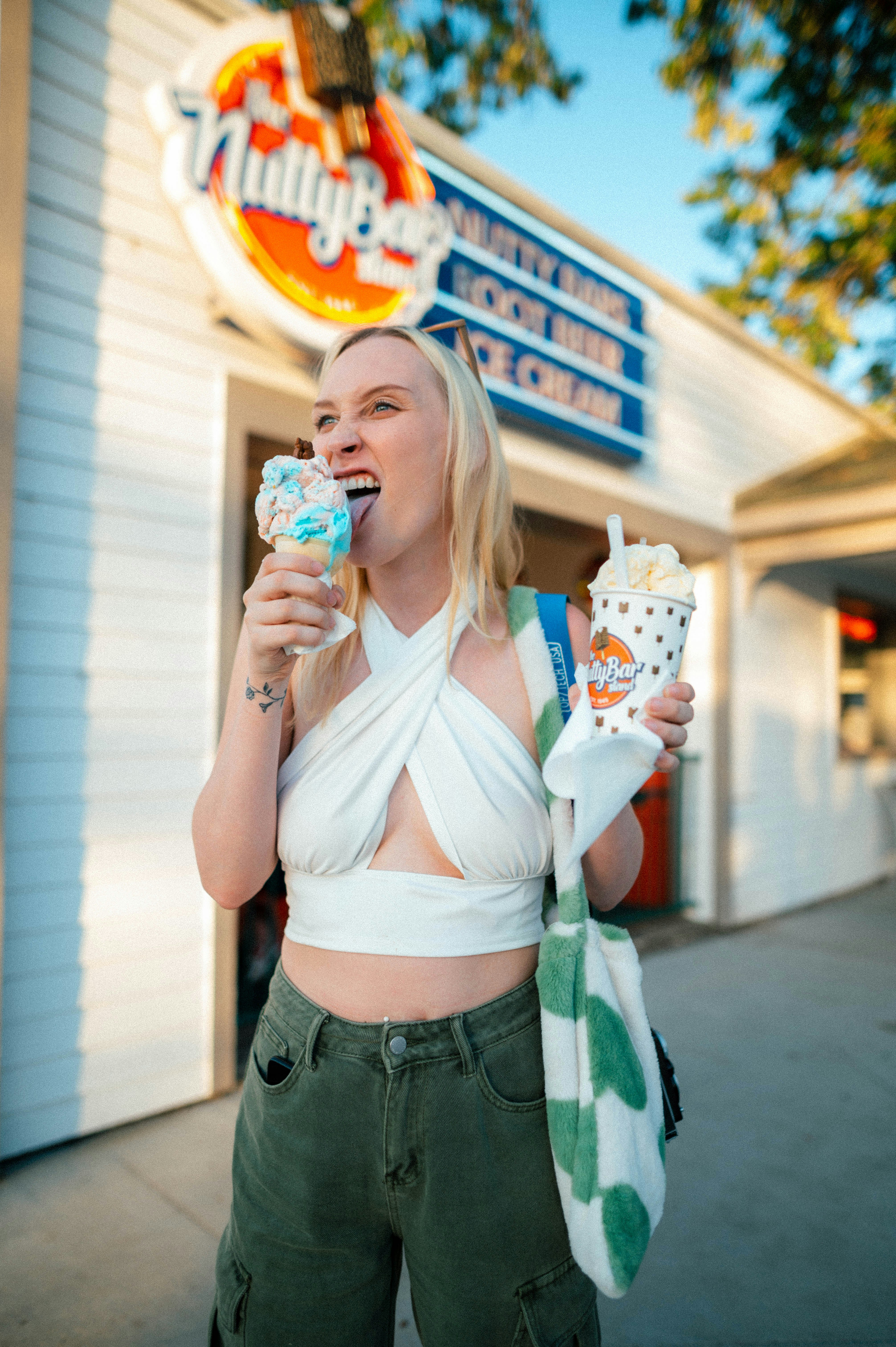 A young woman outdoors enjoys colorful ice cream cones in front of a retro storefront; bright daylight and casual summer attire set a carefree scene.
