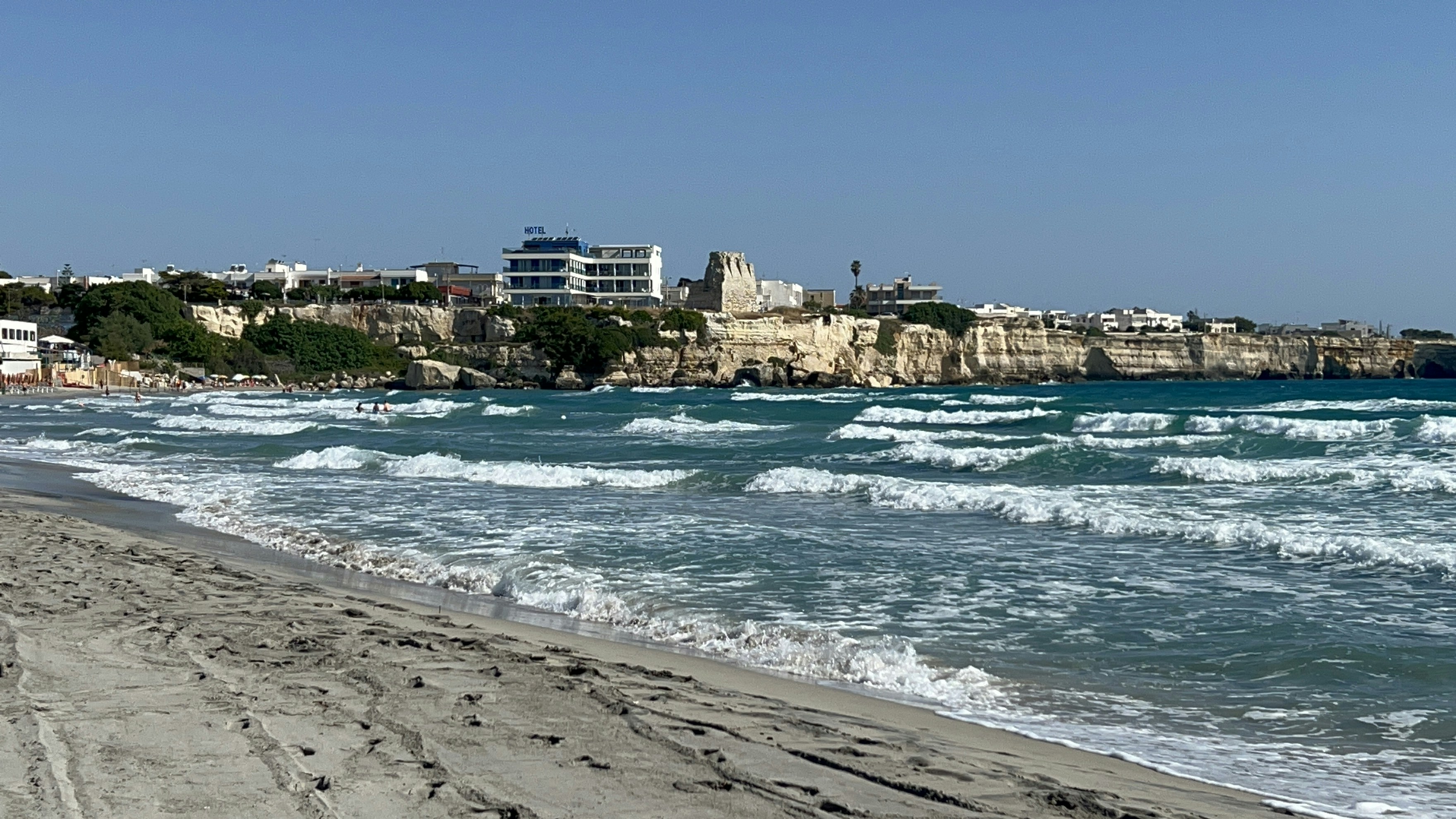 Waves crashing onto a sandy beach with a coastal town and clear blue sky in the background.